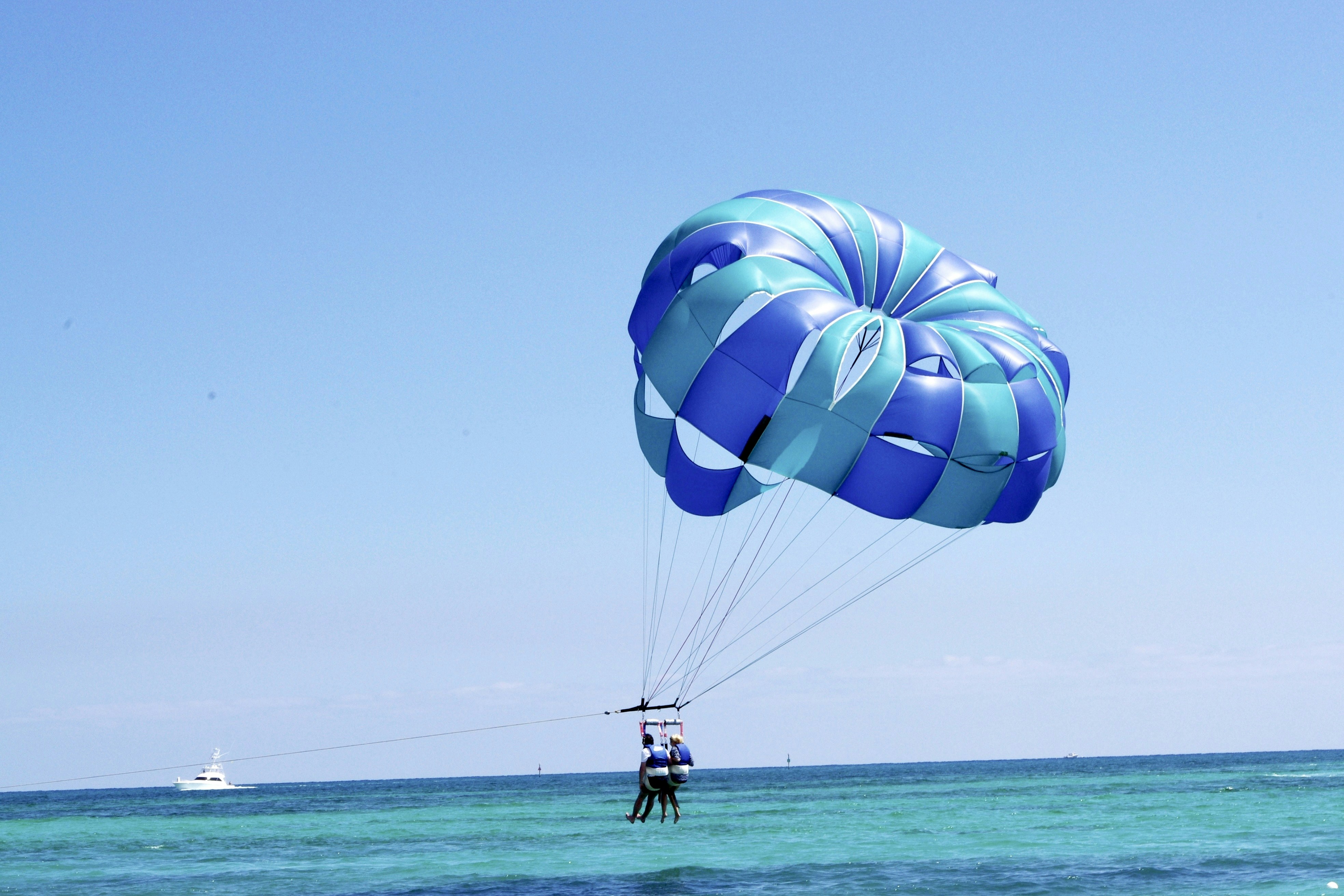 Parasailers hover above turquoise water with a vibrant blue canopy, anchored from a small boat near a calm horizon.