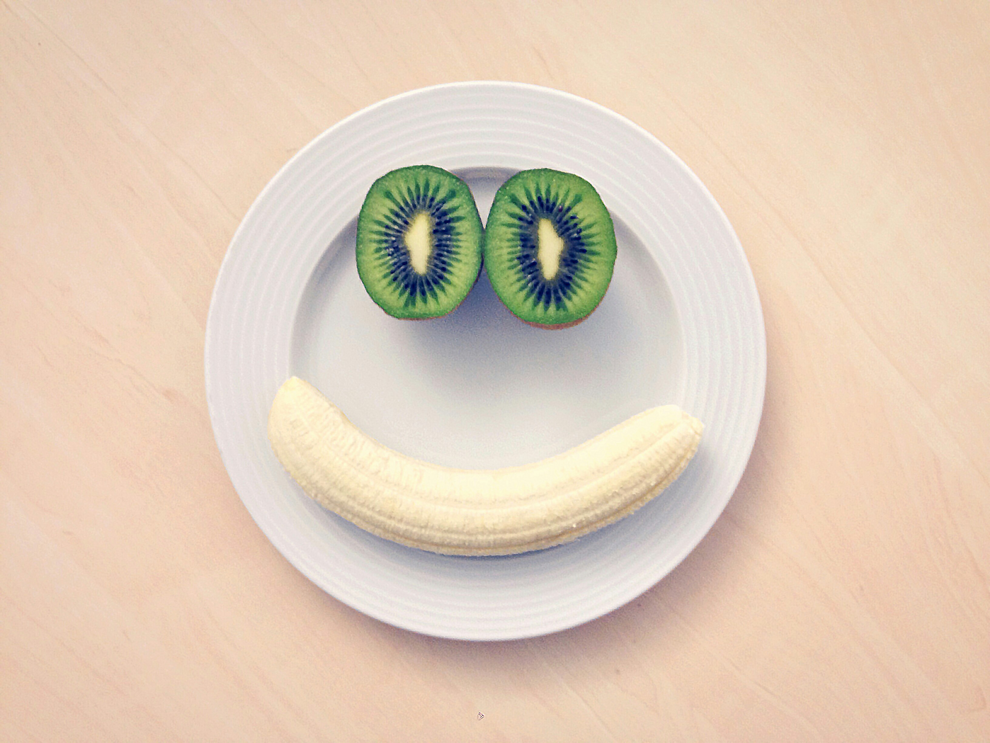 Two kiwi halves form eyes and a peeled banana creates a cheerful smile on a white plate.