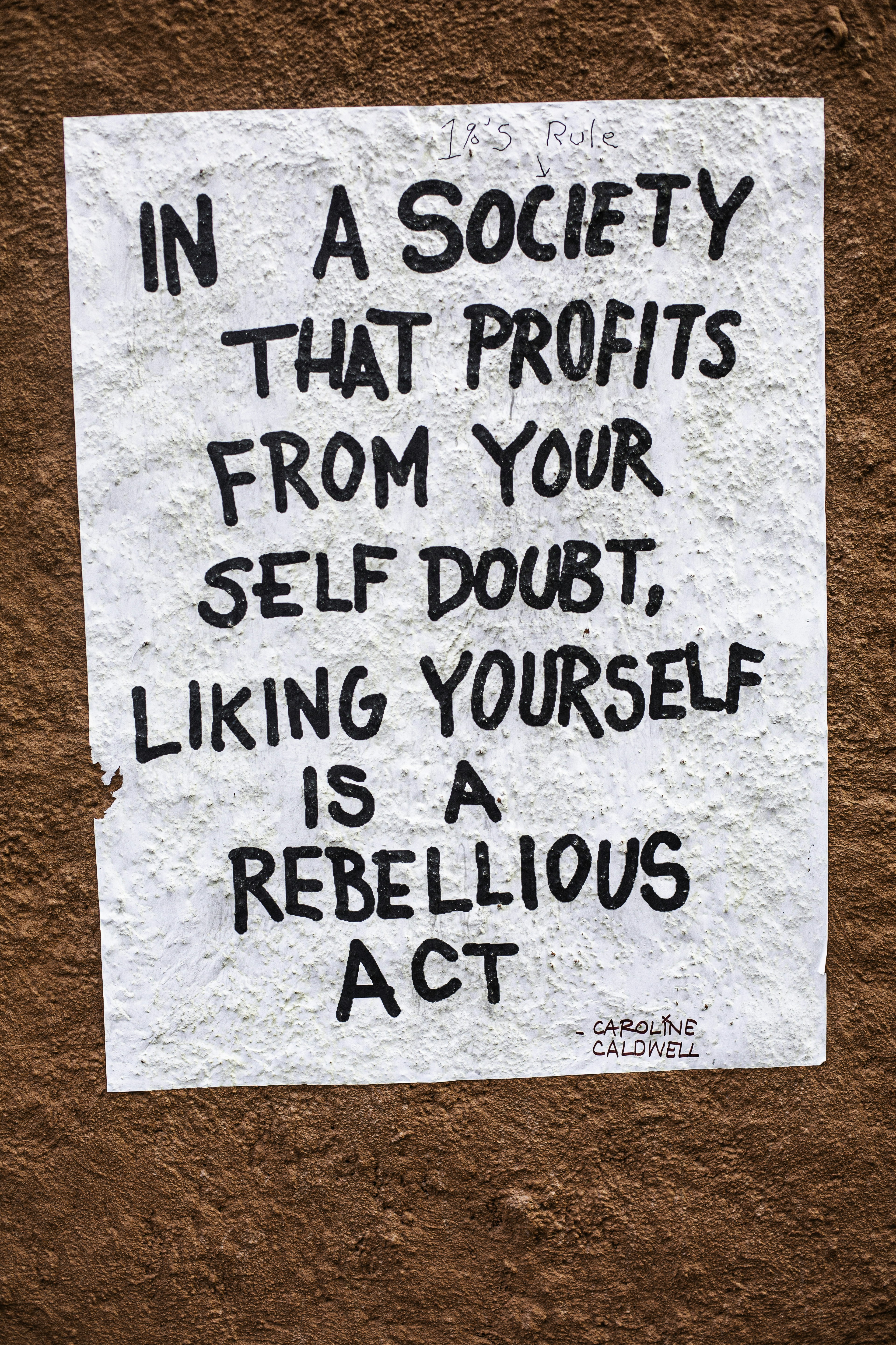 A white quote poster pasted on a rough reddish-brown wall reads “IN A SOCIETY THAT PROFITS FROM YOUR SELF DOUBT, LIKING YOURSELF IS A REBELLIOUS ACT - CAROLINE CALDWELL,” with a small handwritten note above it that appears to read “1%’s Rule.”