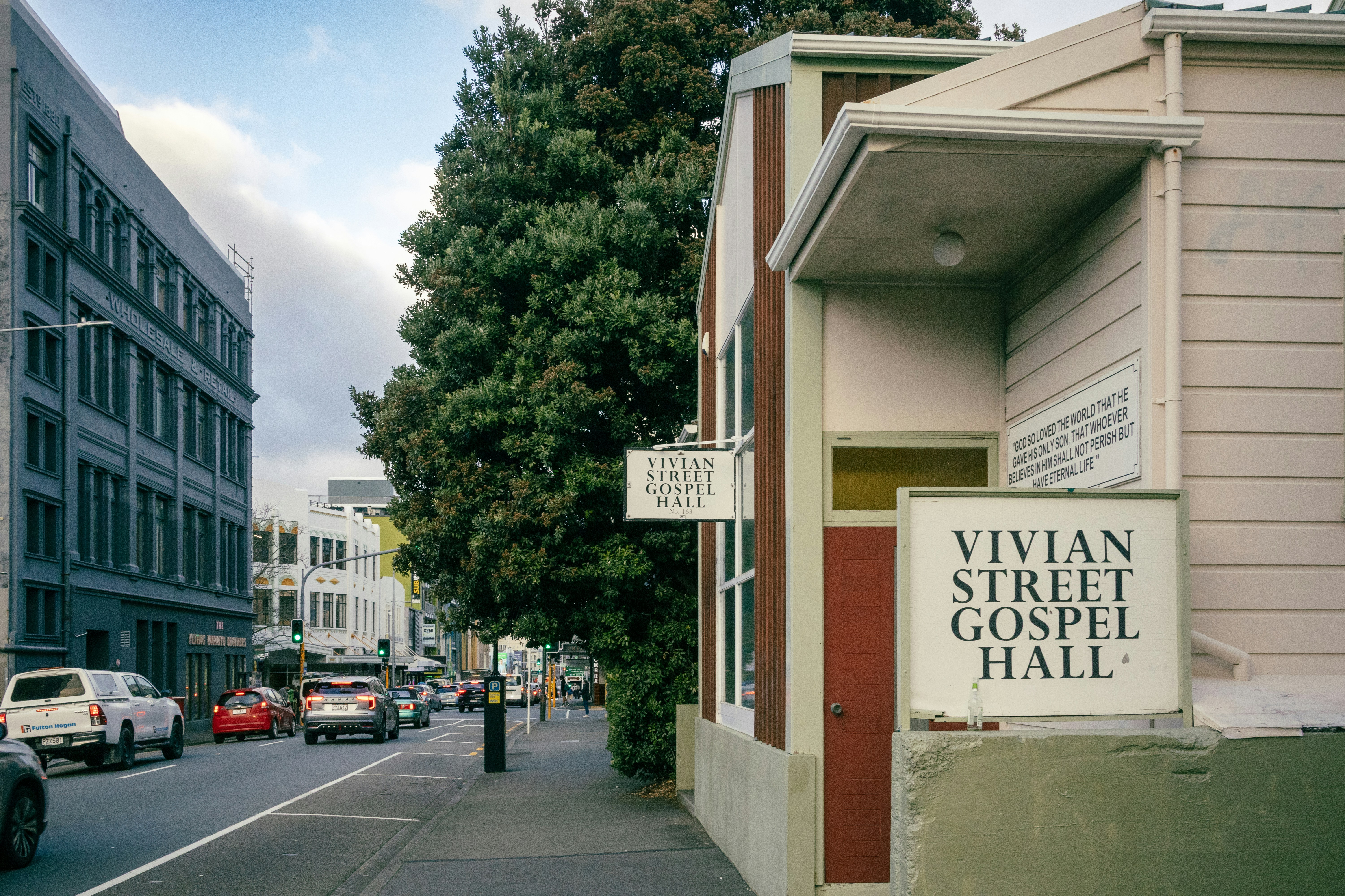 A street view highlights Vivian Street Gospel Hall with a pale façade at right, while traffic and buildings recede along the left. The scene captures an urban corner with contrasting architectural styles and a tree-lined background.
