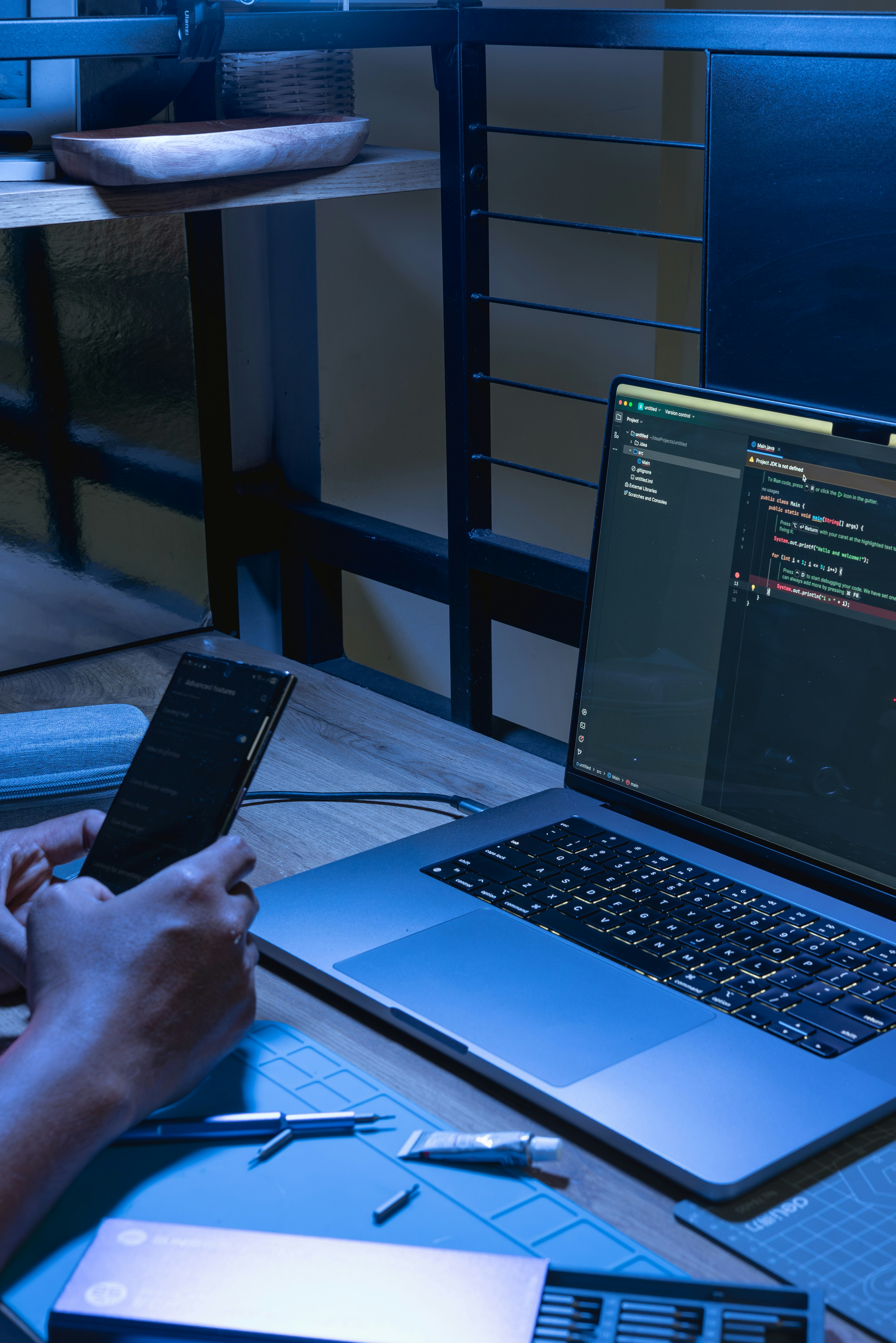 Dimly lit desk scene with a laptop displaying code and a person holding a smartphone; blue-tinted lighting highlights tools and a keyboard trackpad setup, conveying a focused debugging session.