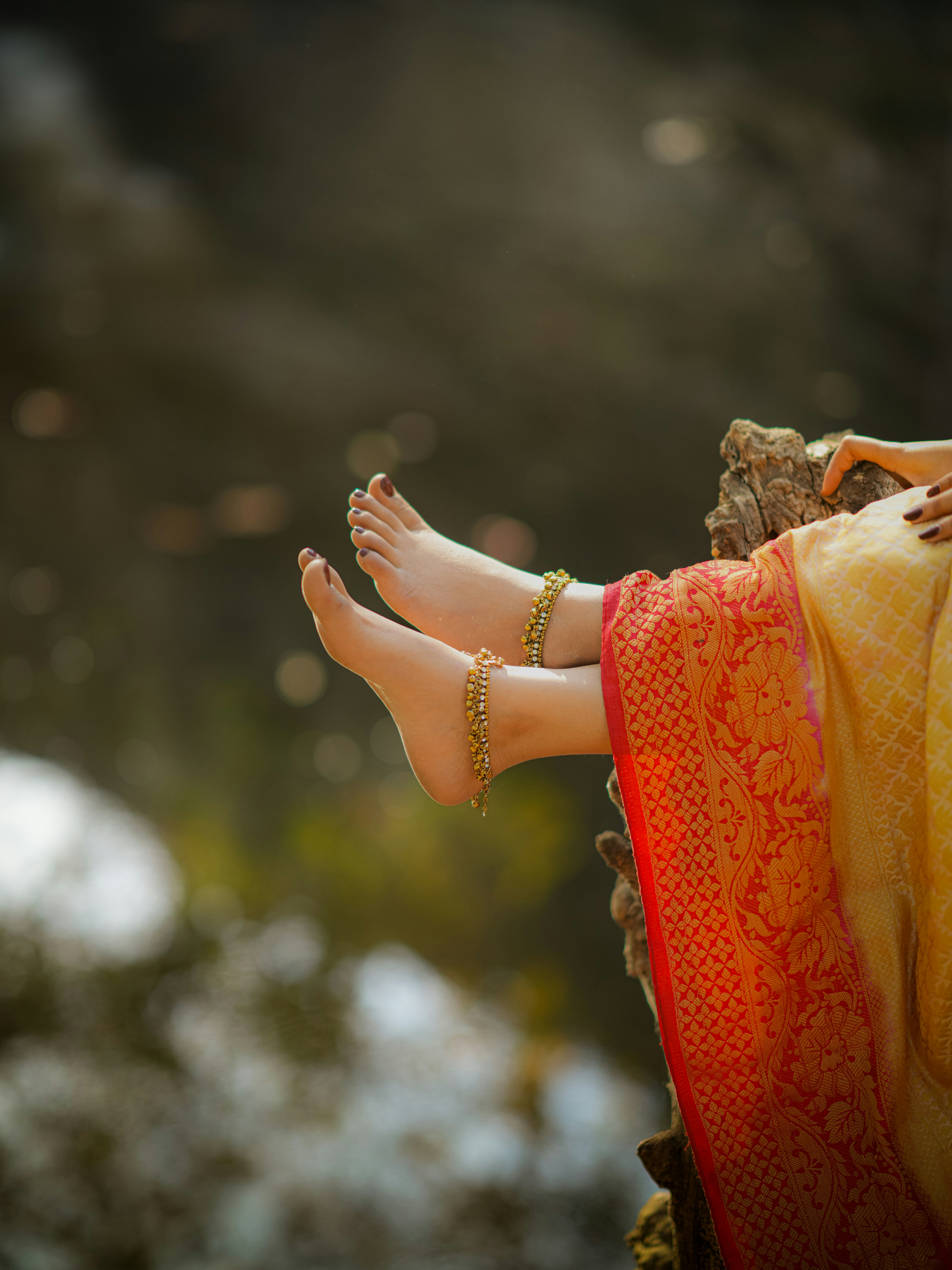 Close-up of feet adorned with gold anklets and a vibrant orange-yellow sari ending, resting on a rock beside a blurred natural backdrop.