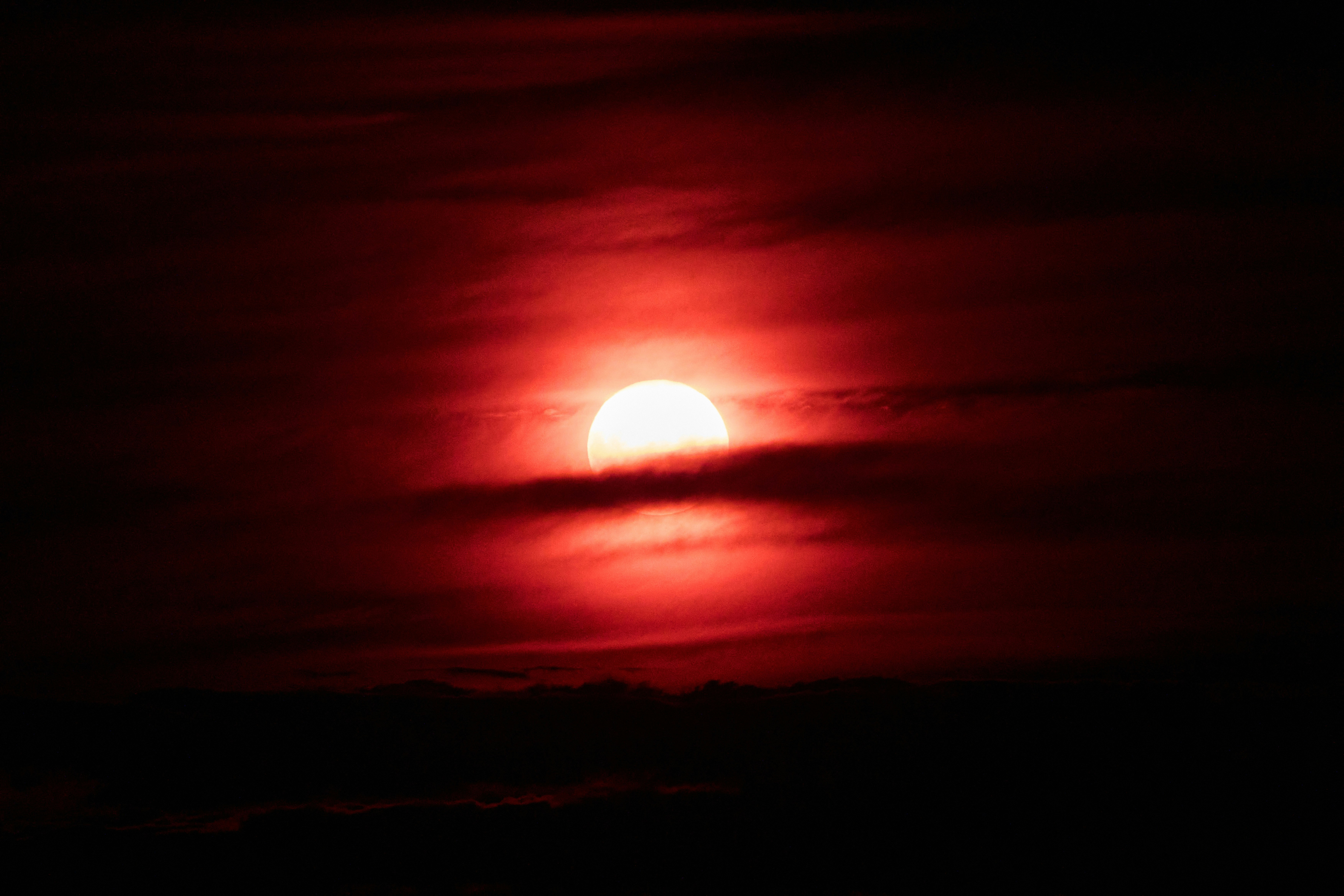 Deep red sunset peeks over dark clouds, with a glowing sun just above a silhouetted horizon. A moody, atmospheric capture emphasizing color and contrast in a low-light scene.