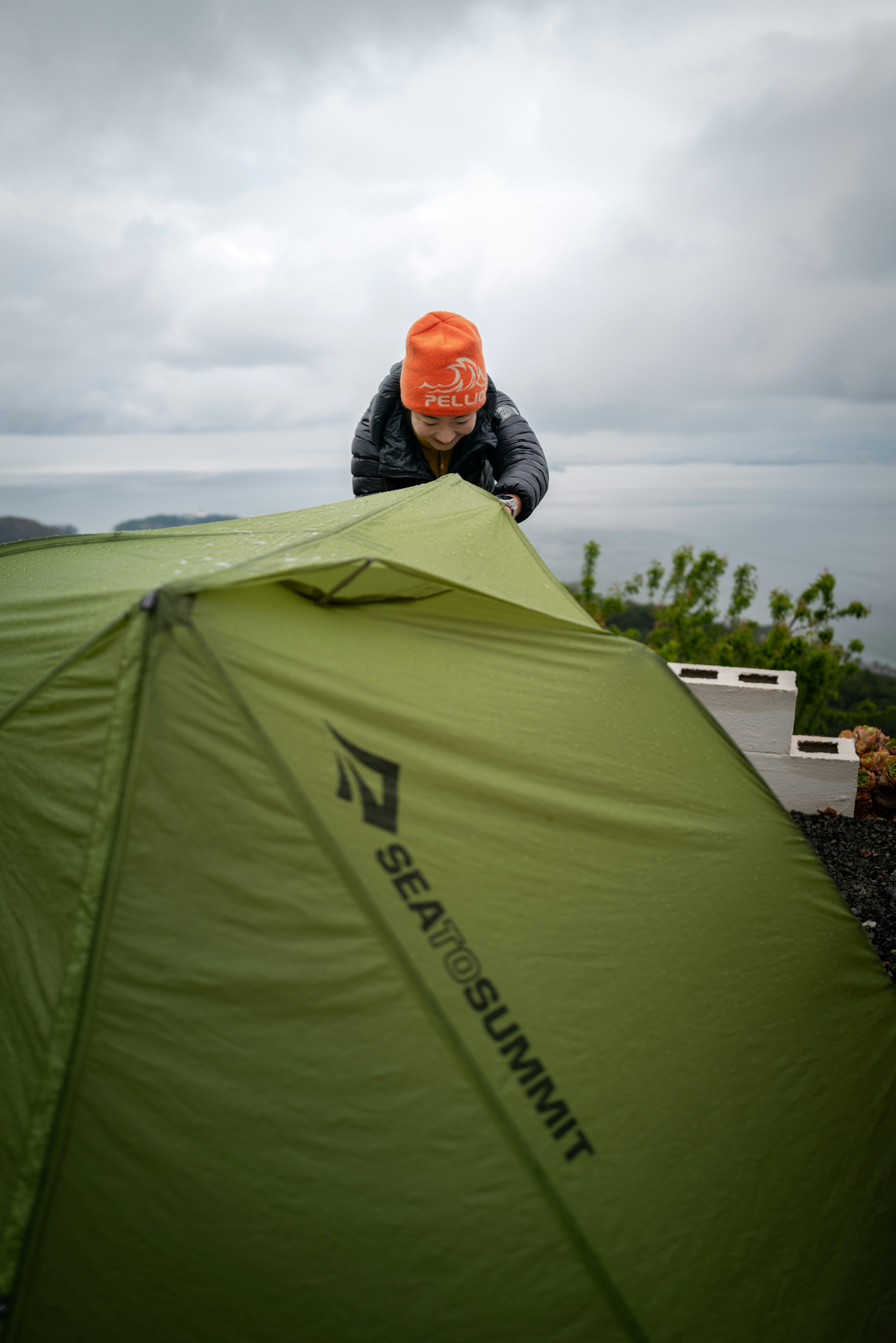 Climber Grips a Green Tent Against a Misty Coastal Sky