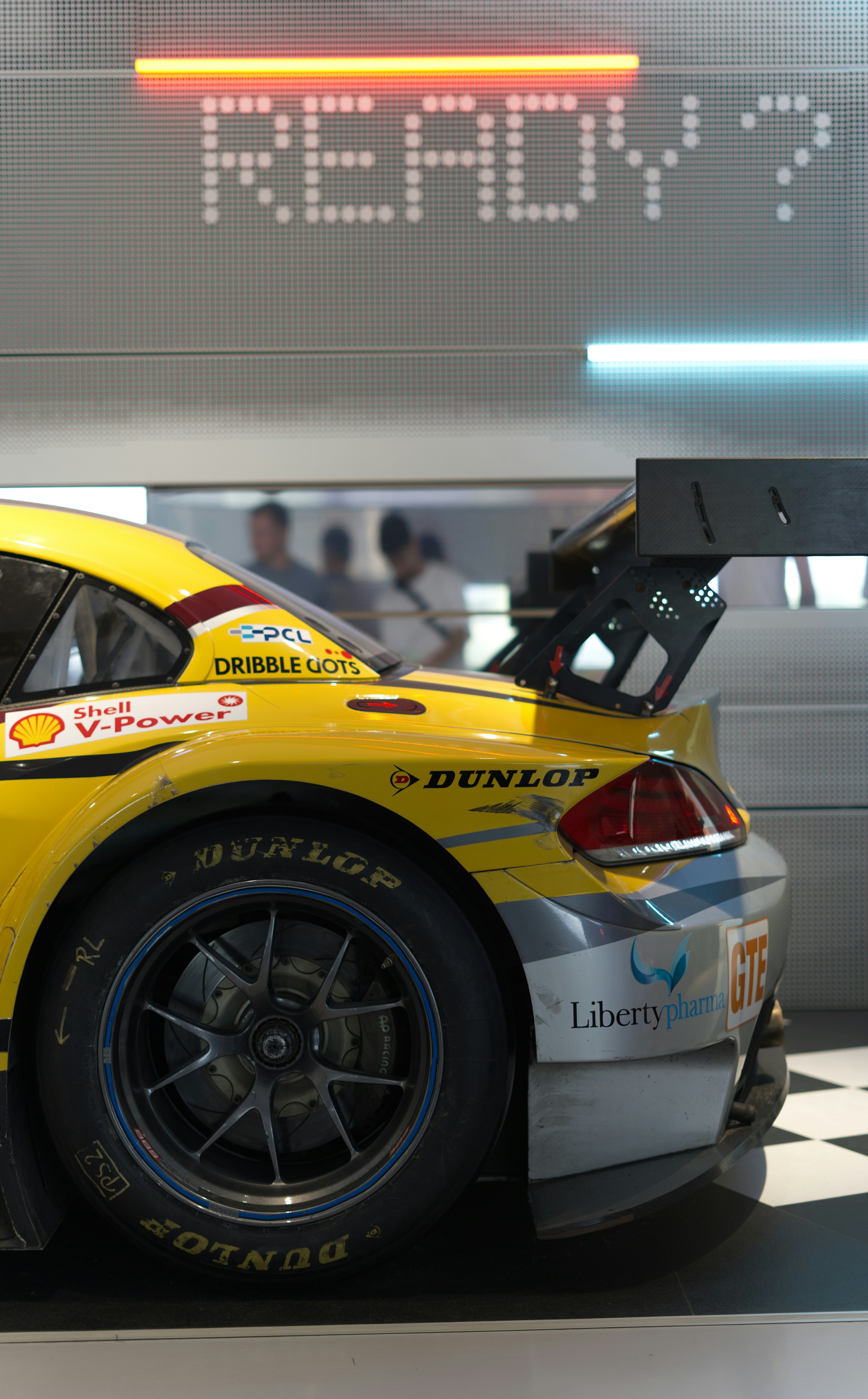 Yellow race car on display with sponsor logos and a checkered floor, captured in a garage-like space with LED signage above.