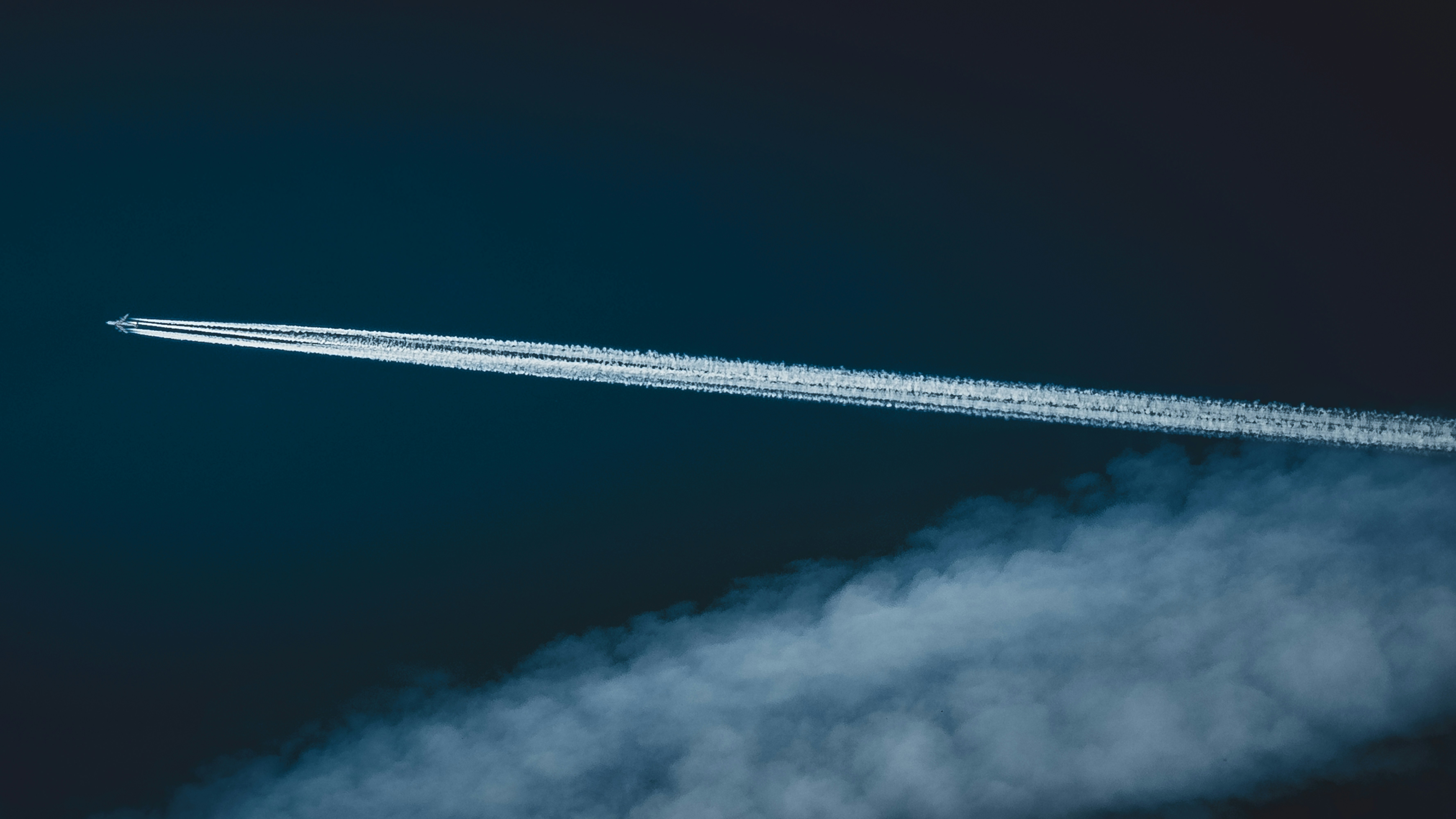 Lone airplane trailing long contrails across deep blue sky above soft clouds.