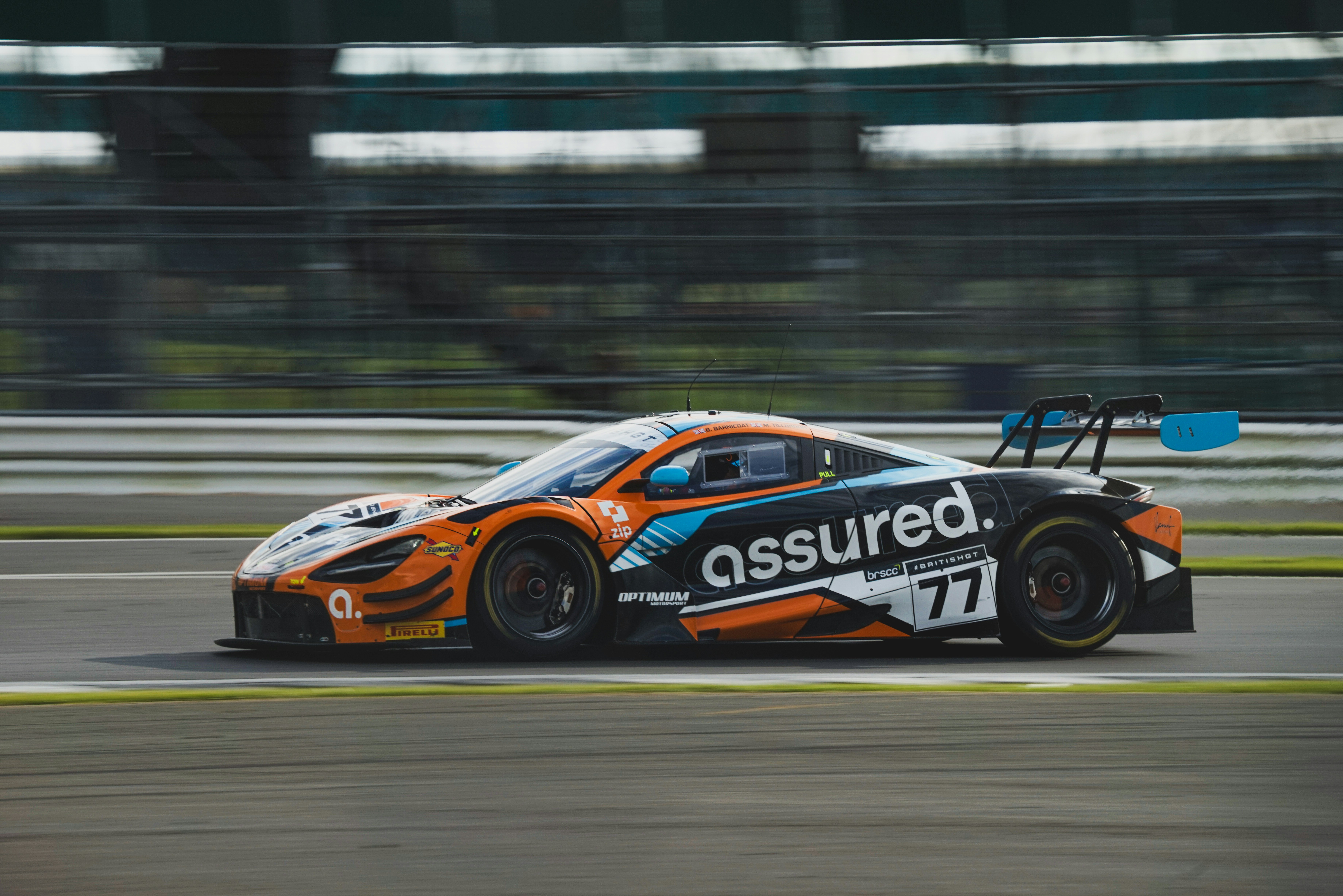 Orange-and-black race car with Assured. branding speeds along a track; the vehicle dominates the frame with a blurred background to emphasize motion and speed.