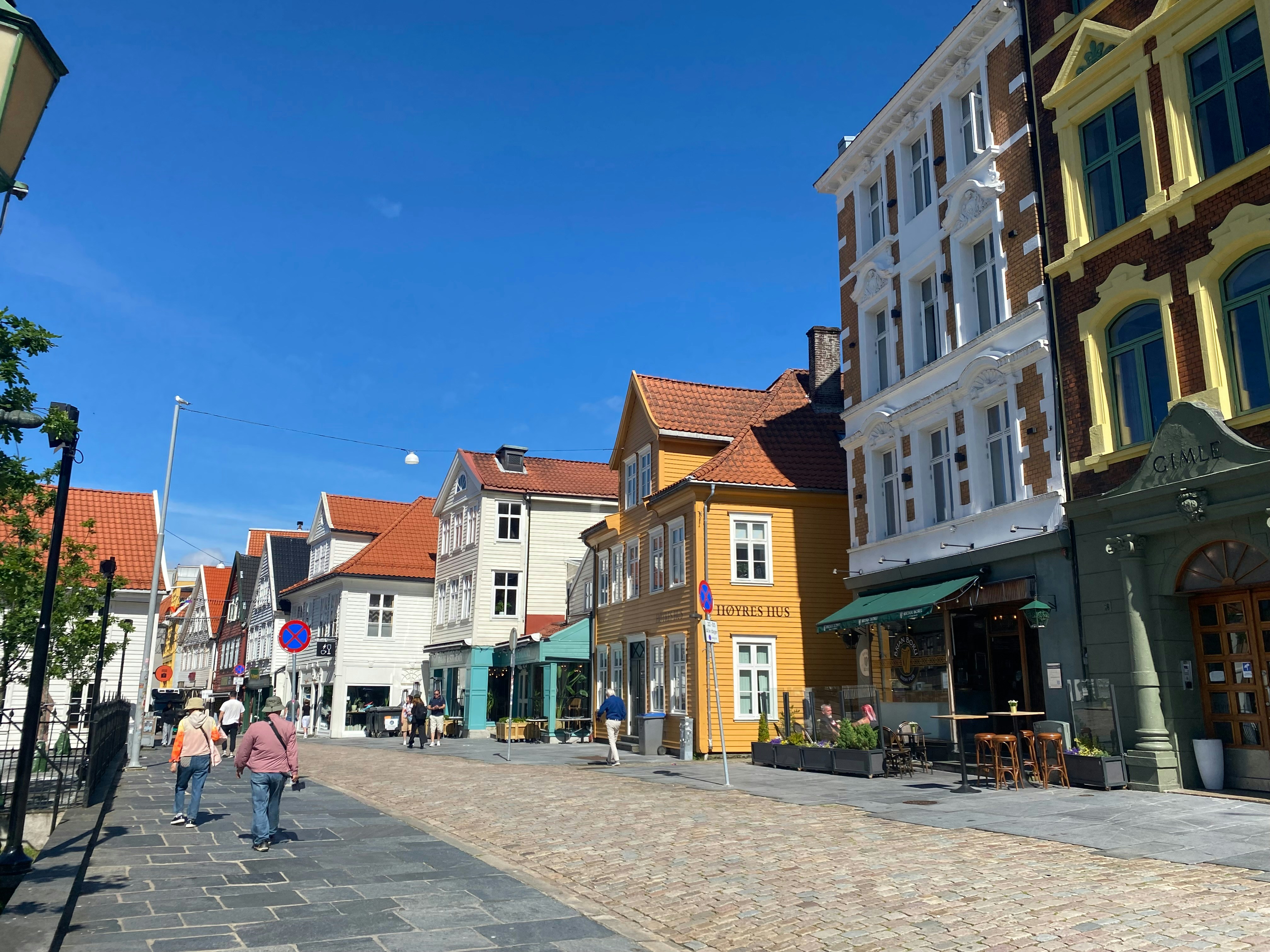 A lively cobblestone street lined with pastel townhouses and outdoor cafés, capturing pedestrians and a sunny urban scene.