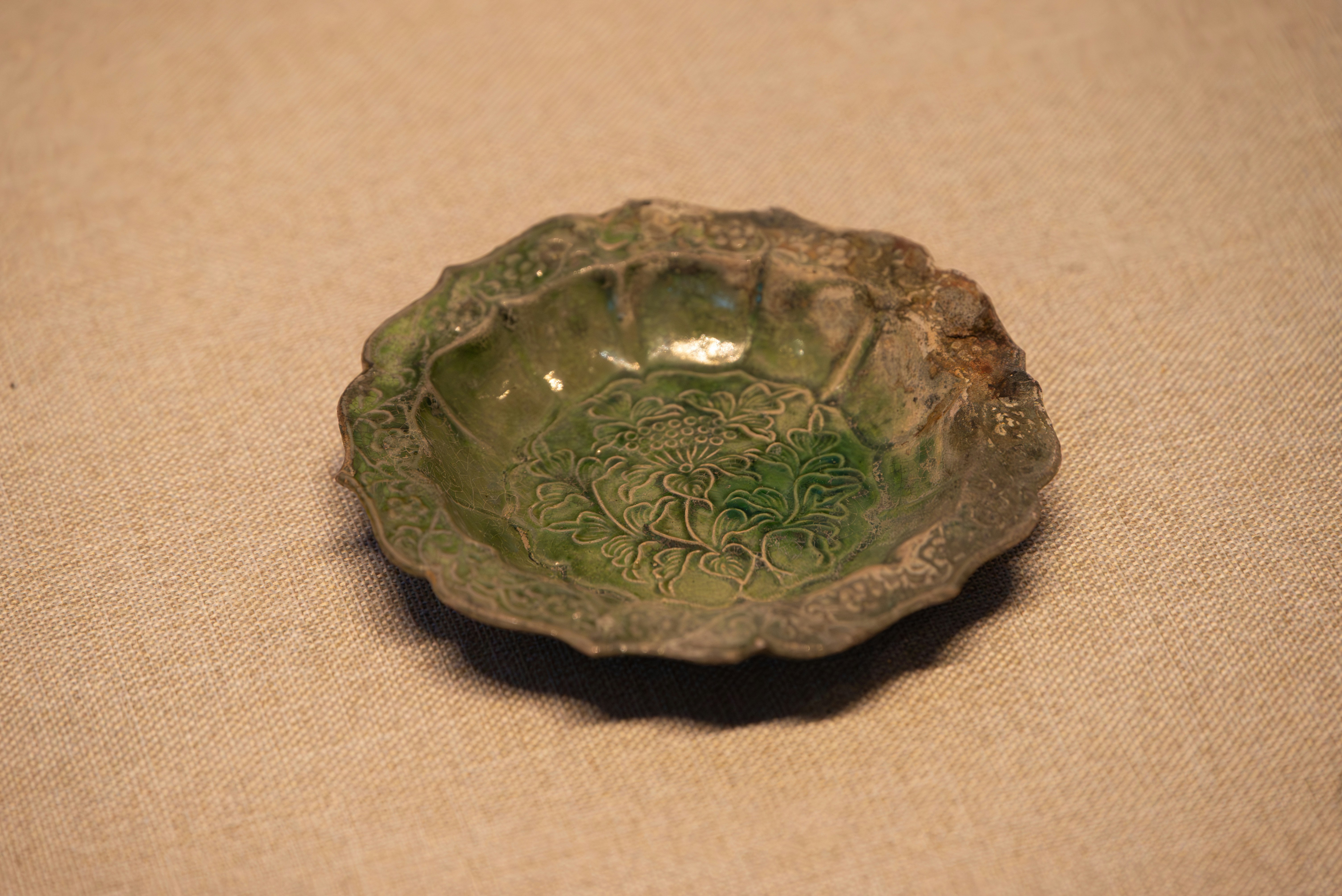 Green ceramic bowl with floral carving rests on beige linen surface.