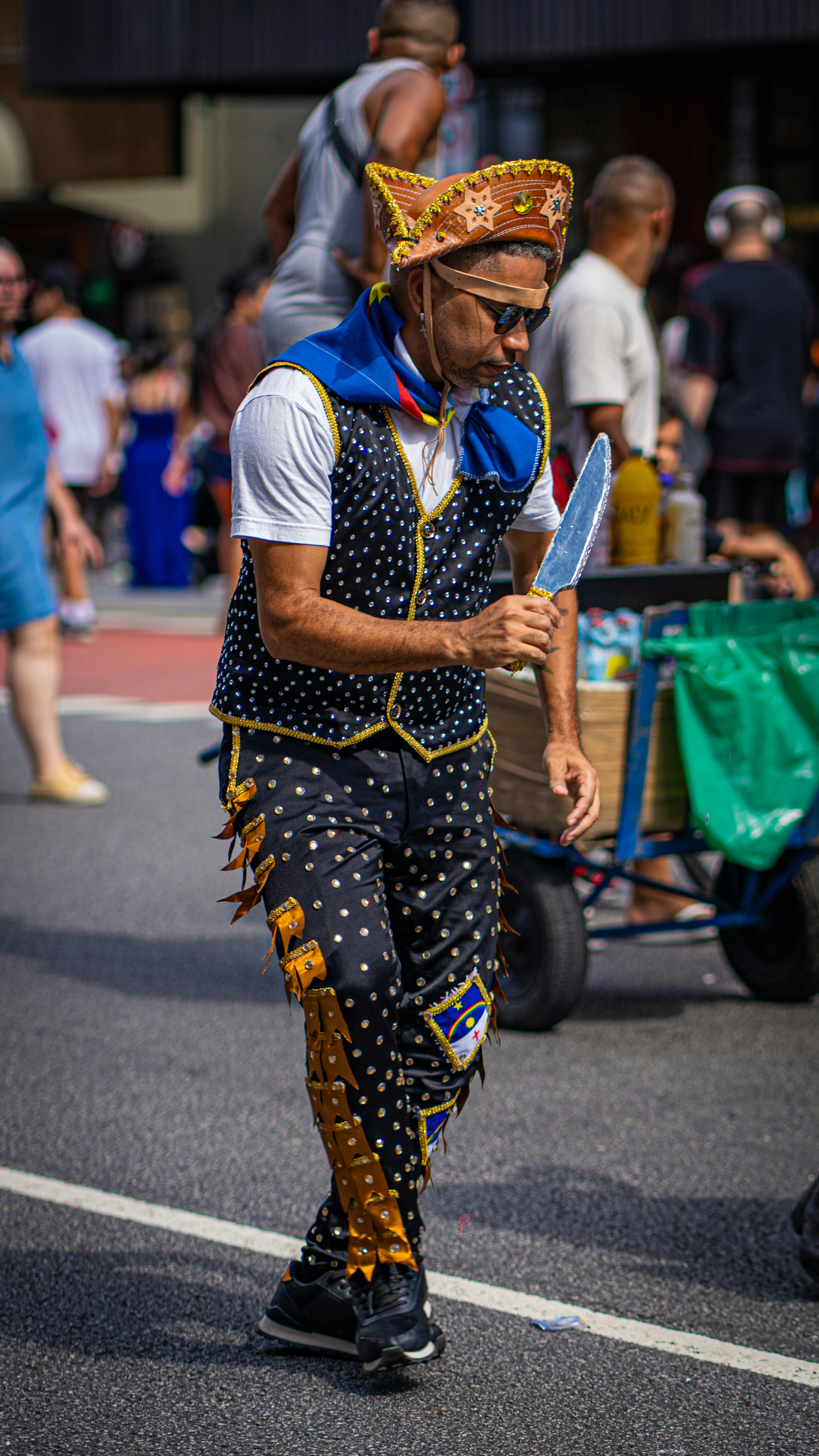Colorful performer in a sequined costume and gold headpiece holds a decorative blade during a street event. Active crowd and vendor setup provide context for a parade-like scene.