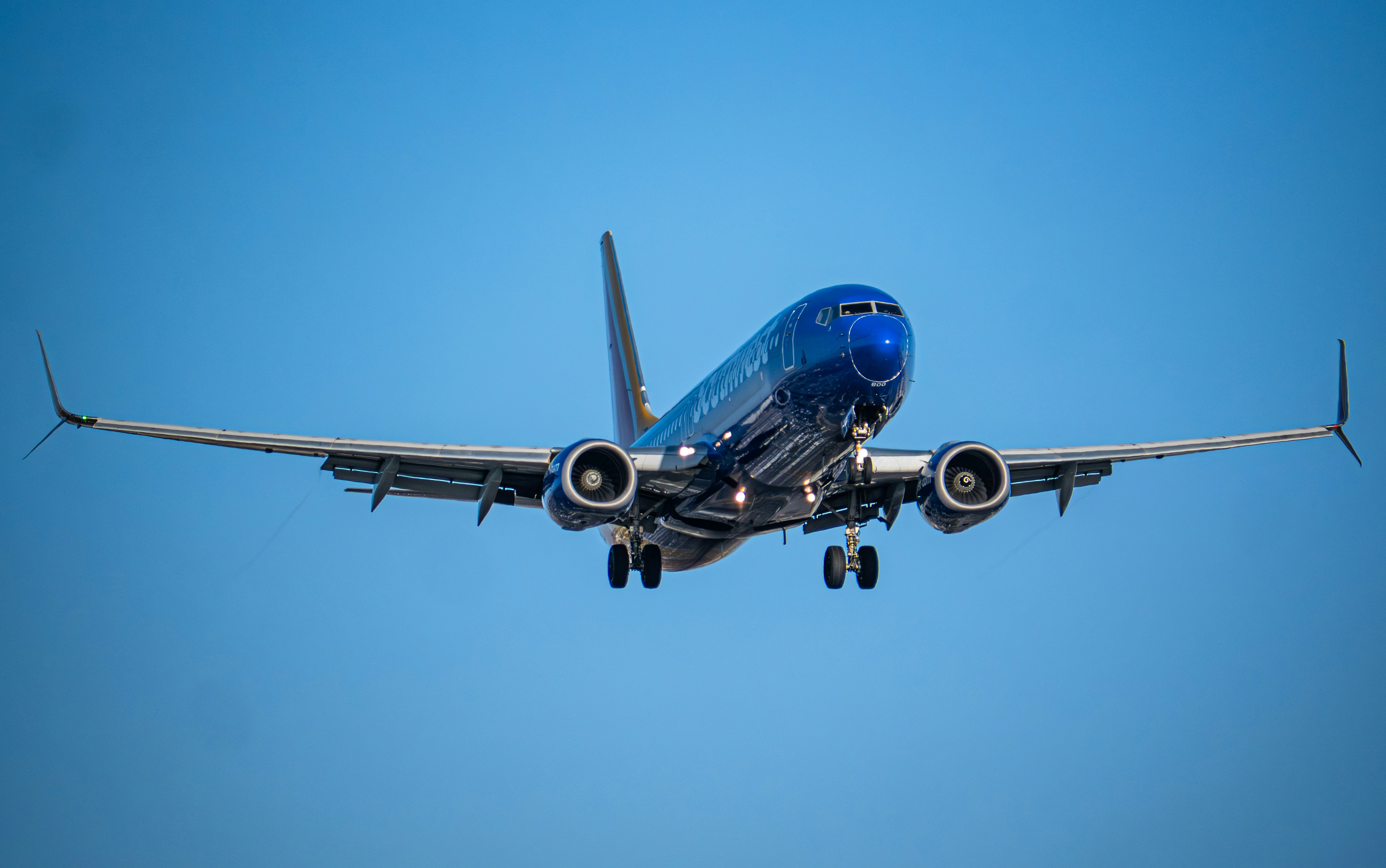 Blue aircraft in mid-approach, nose toward camera with landing gear down against a bright blue sky.