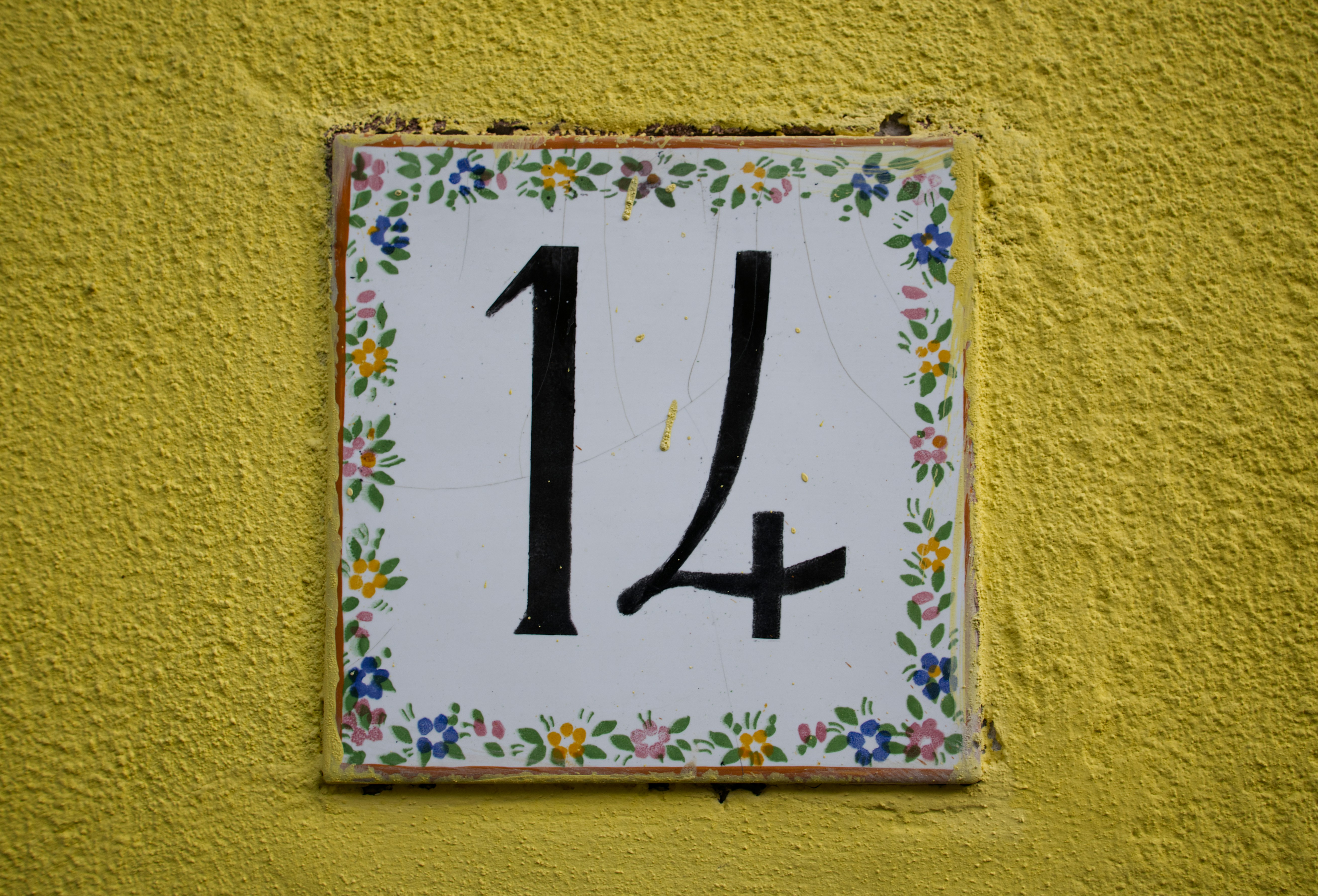 Ceramic Plaque with Floral Border Marks the Number 14 on a Sunlit Yellow Wall