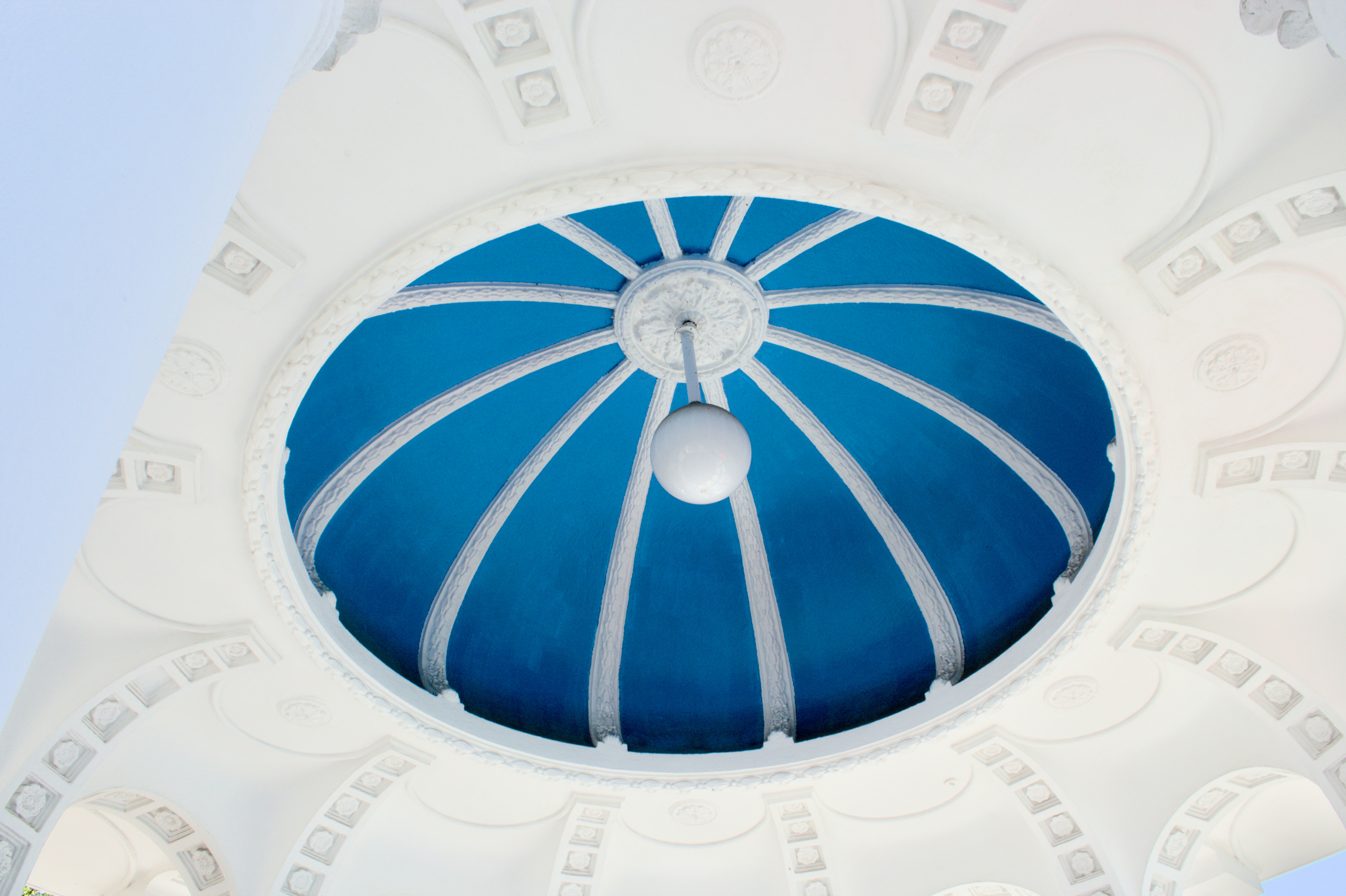 A striking upward view of an ornate architectural dome painted in vivid blue with white radial ribs, set within an elaborately decorated white stone frame. A spherical lamp hangs from the center, emphasizing the geometric precision and classical elegance.