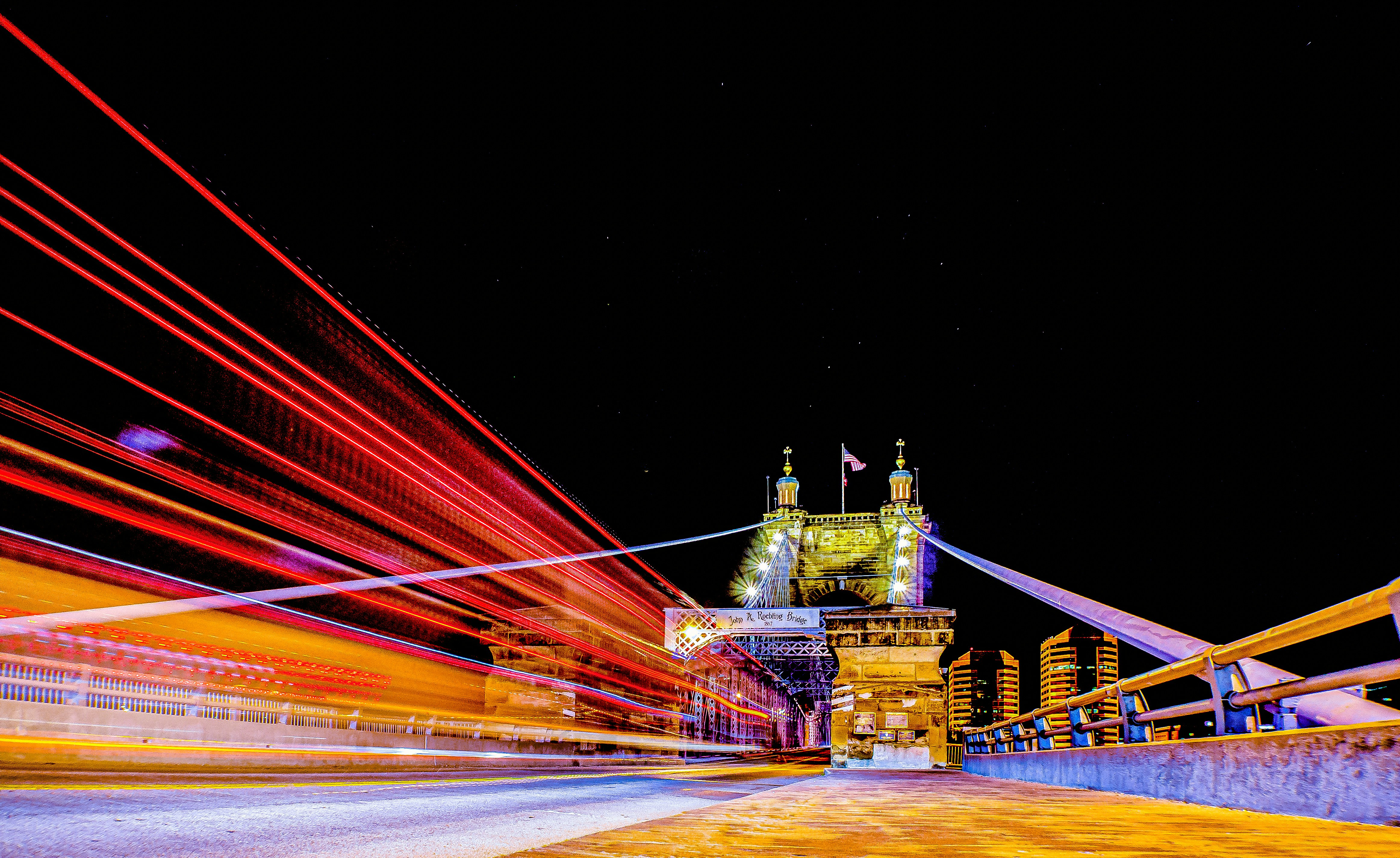 Long-exposure shot of Tower Bridge at night, capturing vibrant light trails from passing traffic across the river surface.