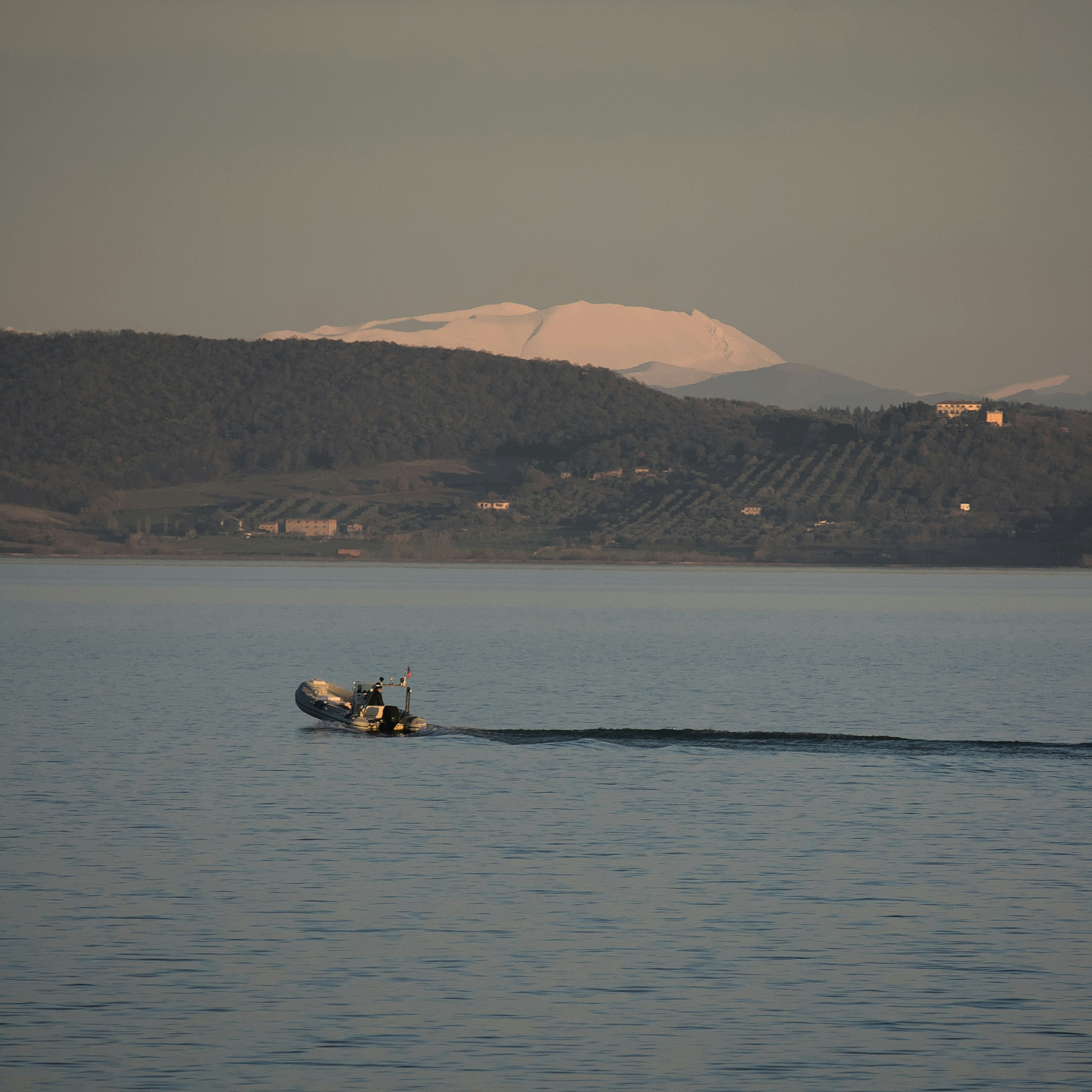 Small boat glides across calm water with distant hills and snow-capped mountains in the background, under soft evening light.