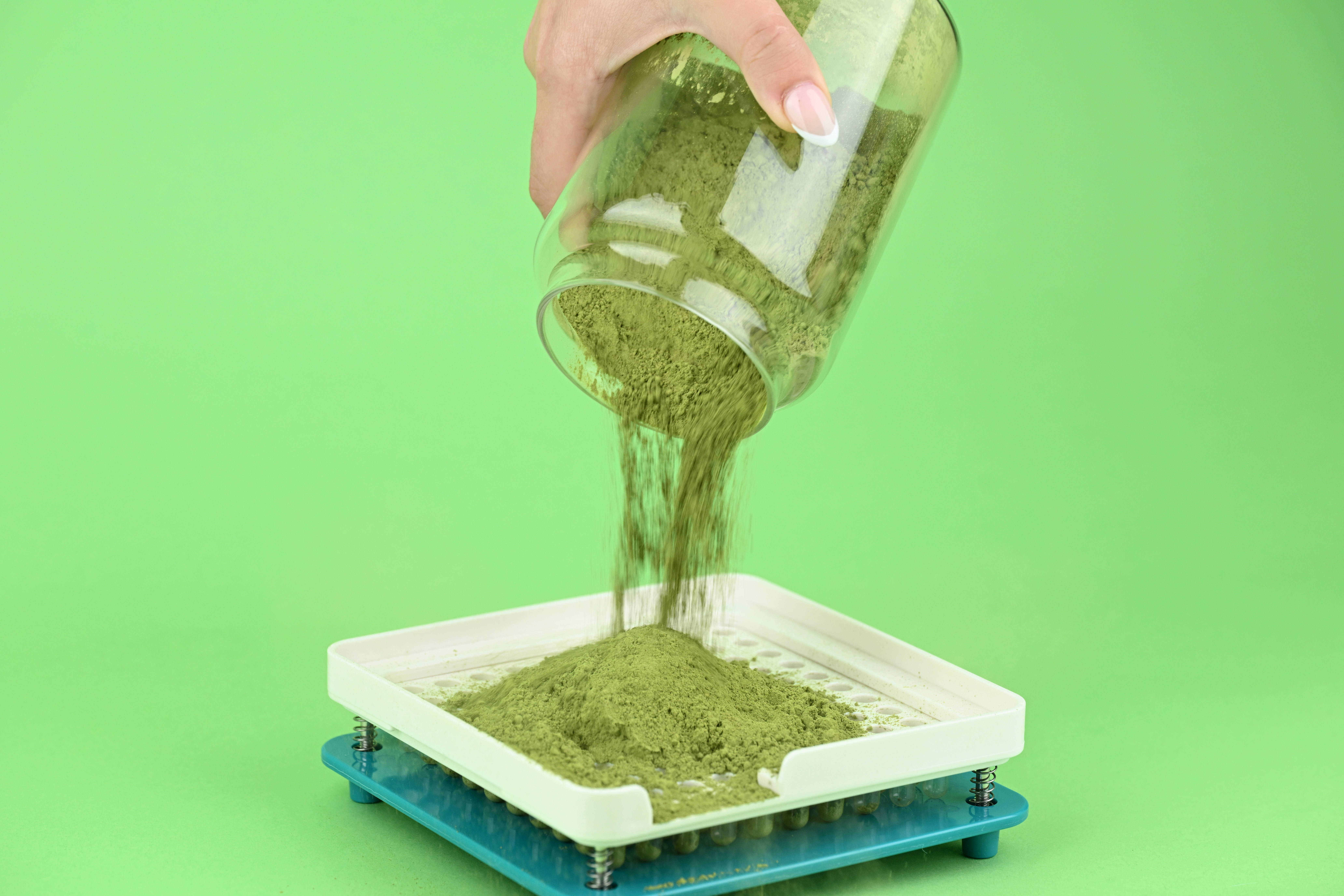 Woman's hand pouring green herbal supplement powder into a Blated Cap & Press Core Series 100 hole capsule filling machine with herbal powder sitting on top waiting to be spread - Photo courtesy of https://blated.com - credit appreciated