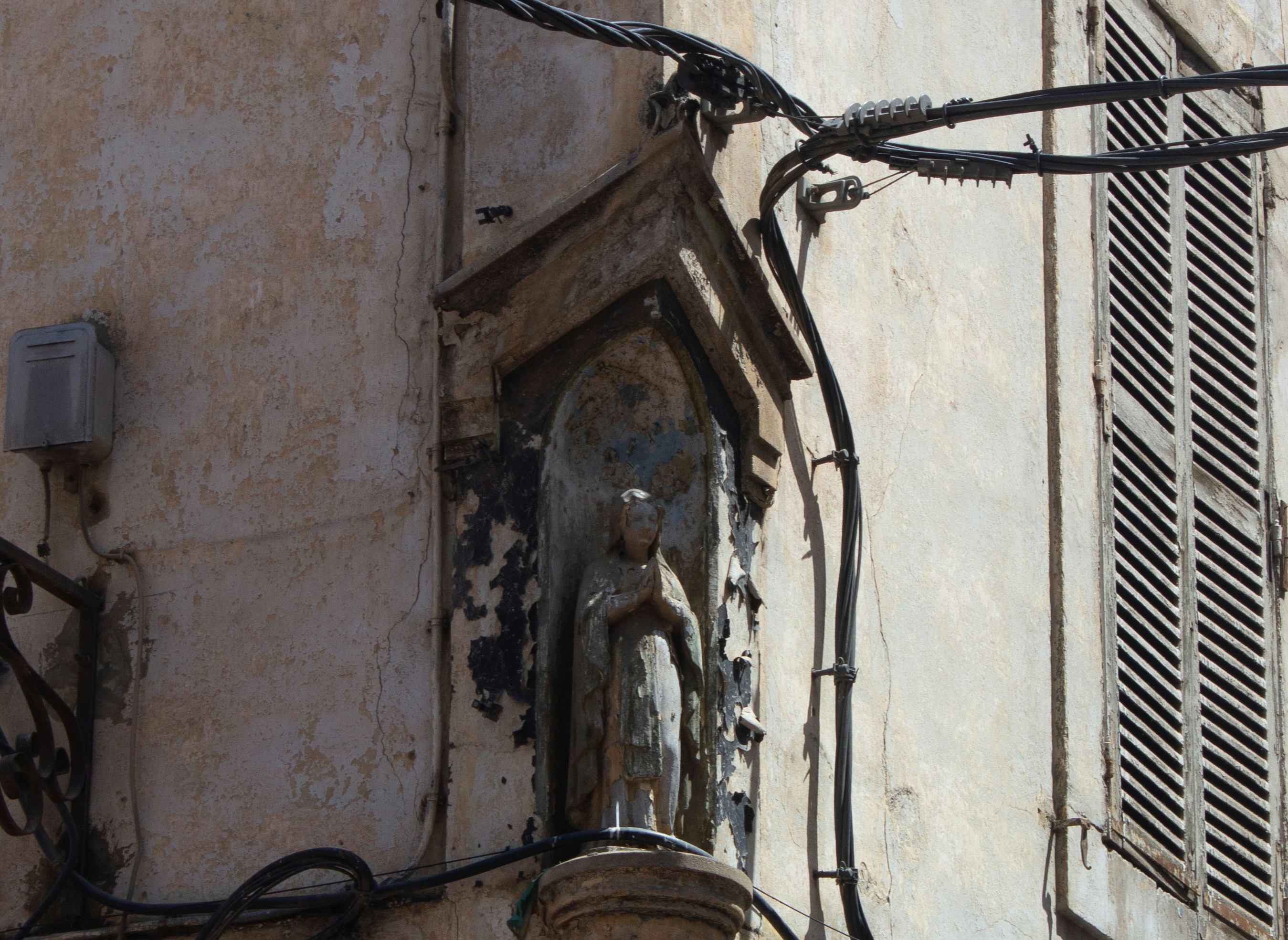 Weathered niche: a solitary statue framed by wires and shutters