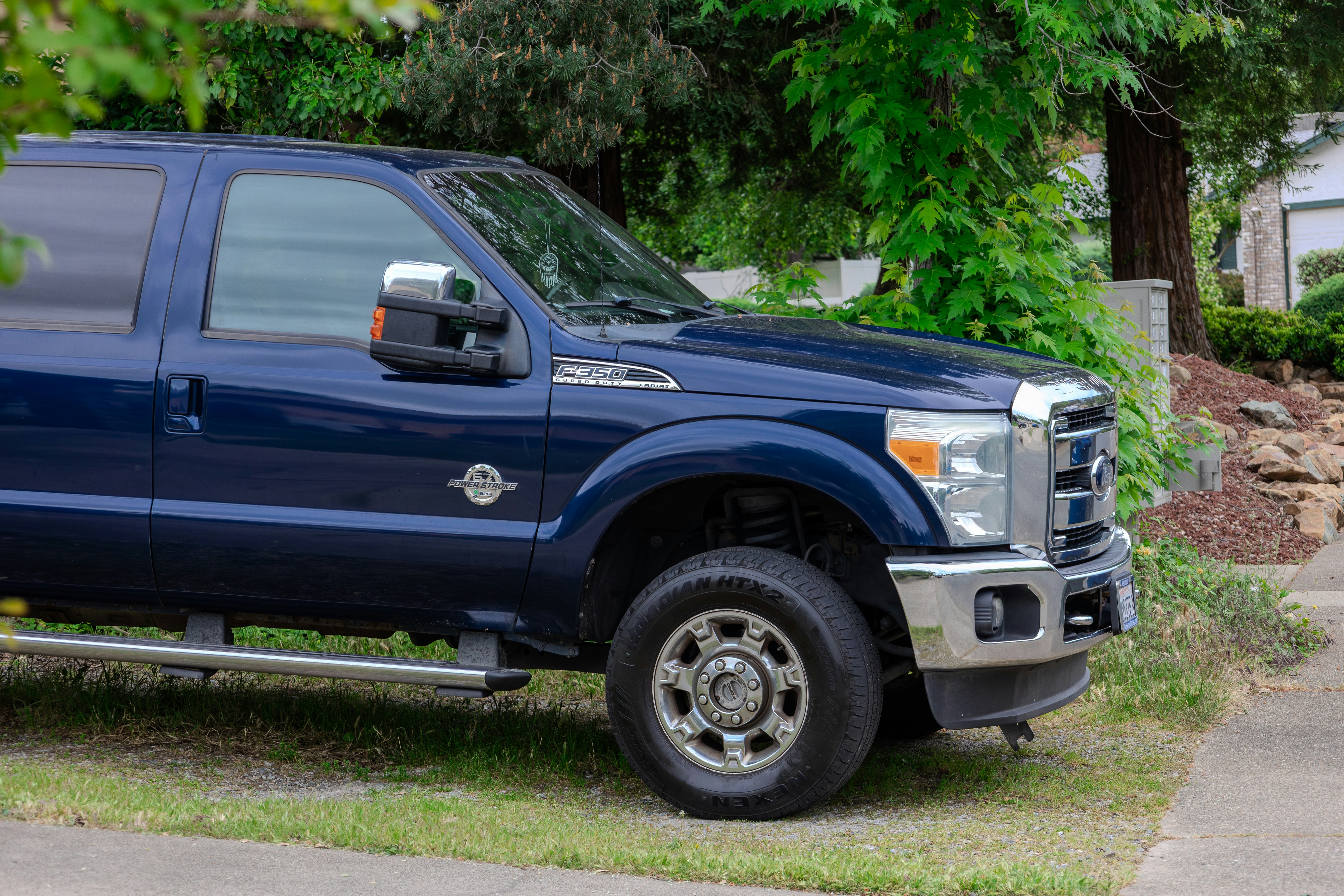 Blue Ford pickup parked on grass by a driveway, with green foliage and a brick landscape in the background. A truck emblem and wheel detail are visible, suggesting a daytime outdoor scene.