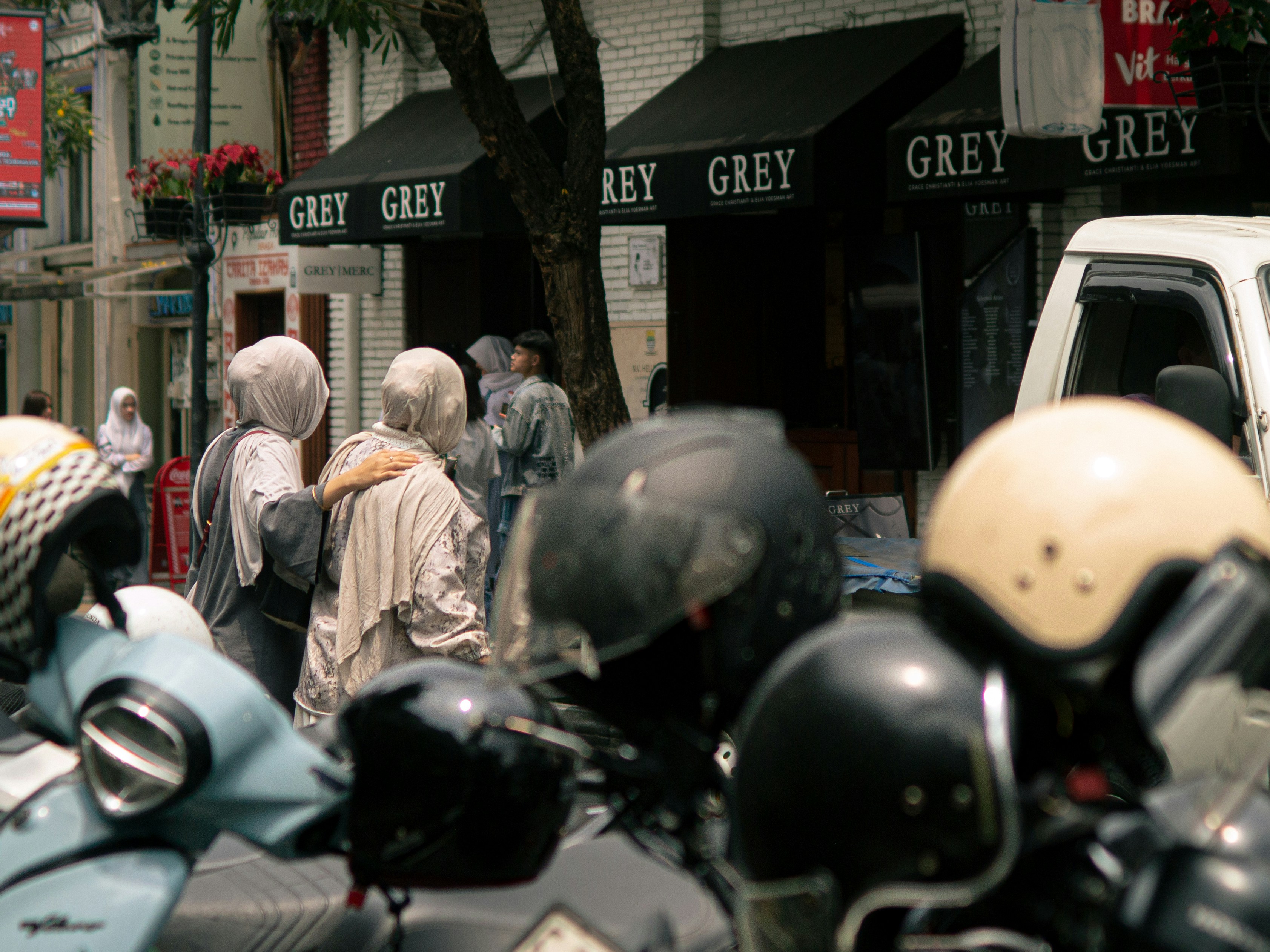Street scene with pedestrians in hijabs behind a row of helmets; storefronts and signage in the background create a busy urban atmosphere.