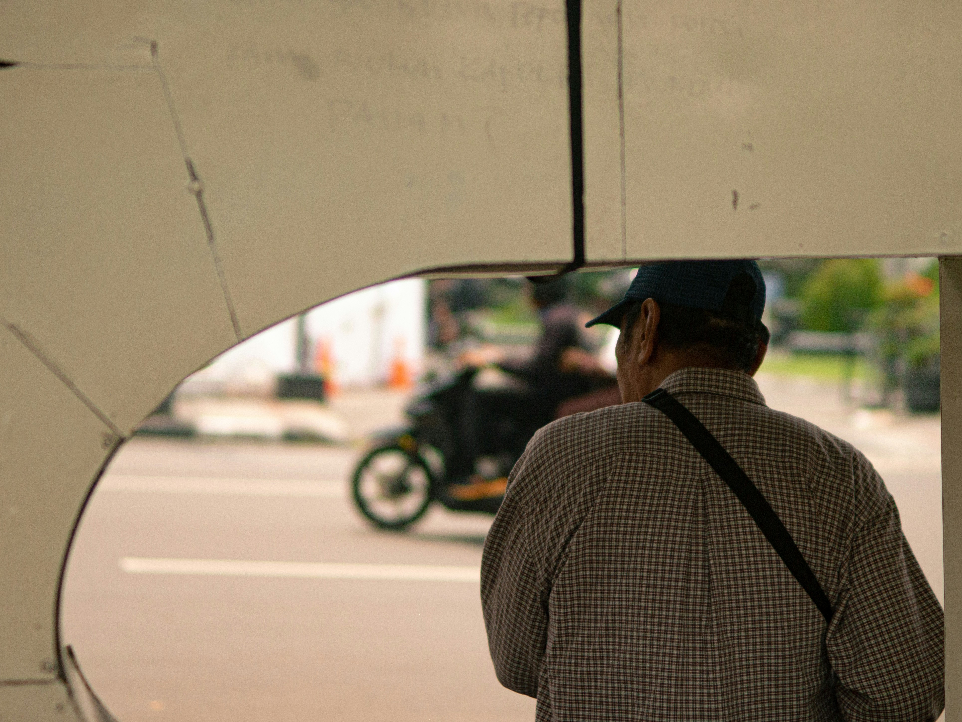 Partial view of a man in a checkered shirt under an architectural arch, with a motorcycle blurred in the background; a candid street scene focusing on everyday detail.