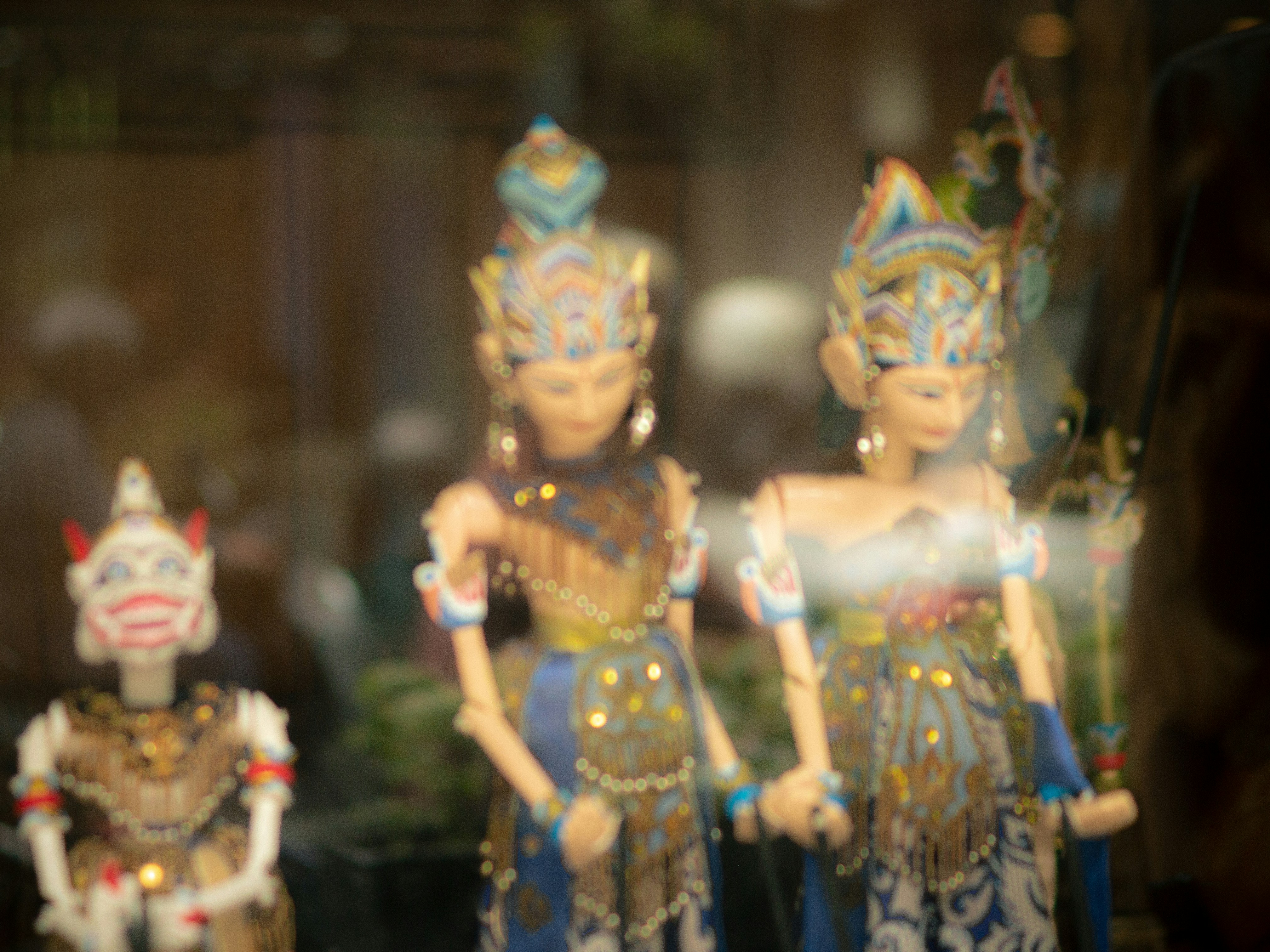 Colorful, ornate dolls behind glass appear blurred, suggesting a storefront display with ambient reflections. The scene emphasizes decorative attire and cultural styling rather than fine facial detail.