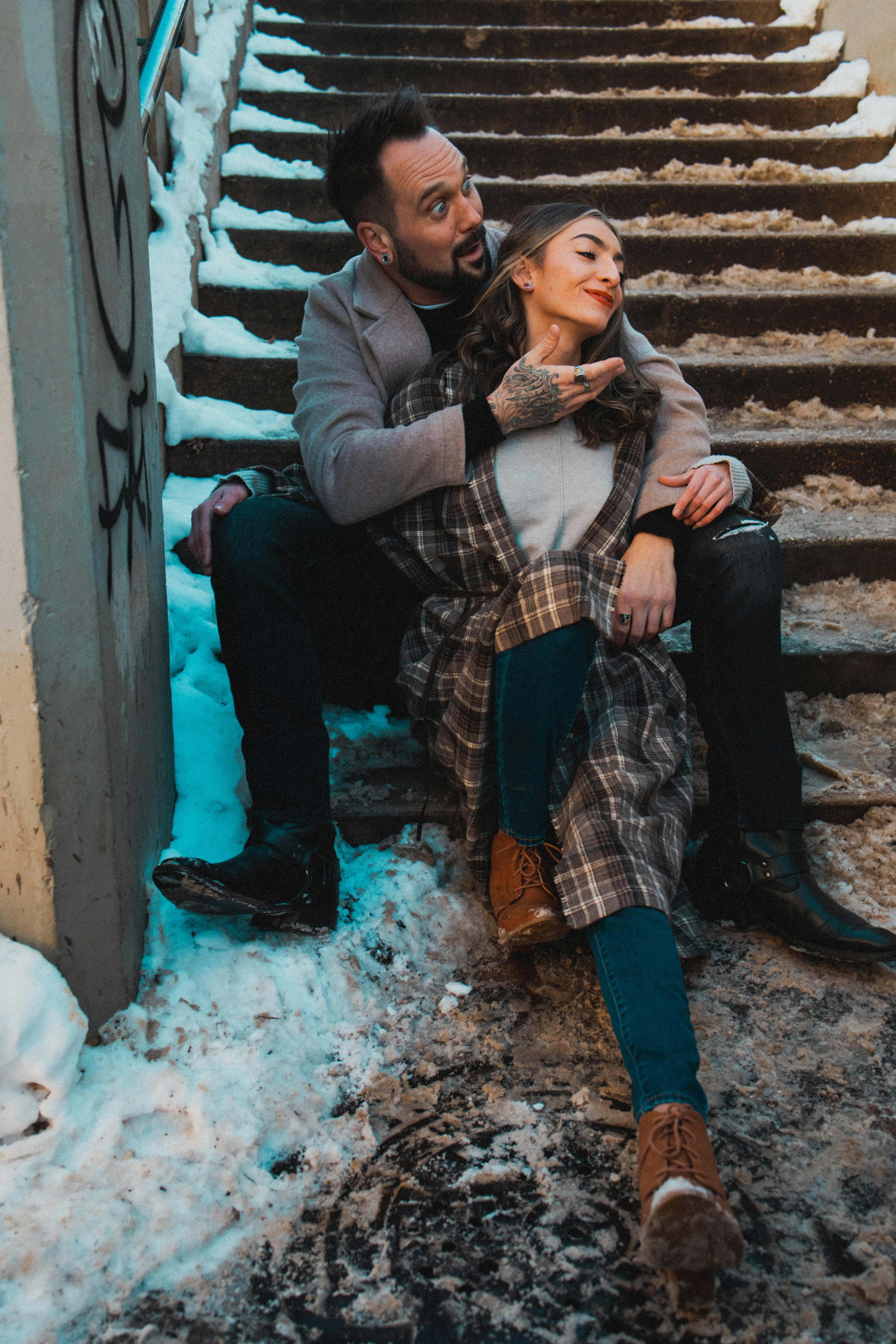 Couple sits close on snowy stairs, the man embracing the woman with a playful gaze as she smiles; urban winter scene with cold tones and warm clothing.
