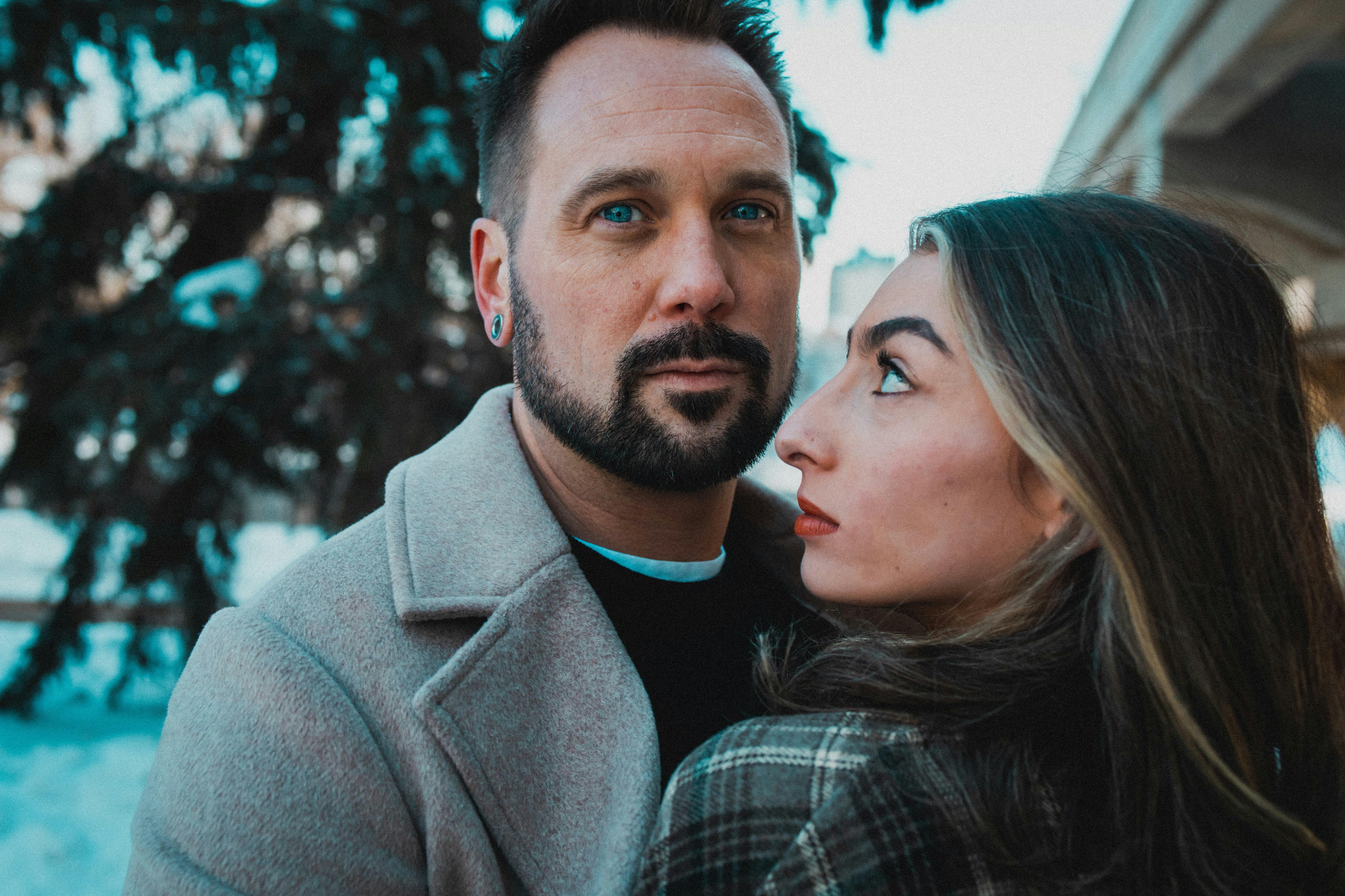 Two people stand close, the man looking past the camera while the woman gazes at him; their faces fill the frame with a shallow depth of field and urban backdrop. A candid portrait featuring a couple in a cool-weather scene; photographic emphasis on connection and mood.