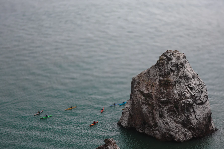 Rock sea stack rising from calm turquoise water with a group of kayakers nearby, capturing a coastal moment. The image emphasizes scale and tranquility of the seascape.