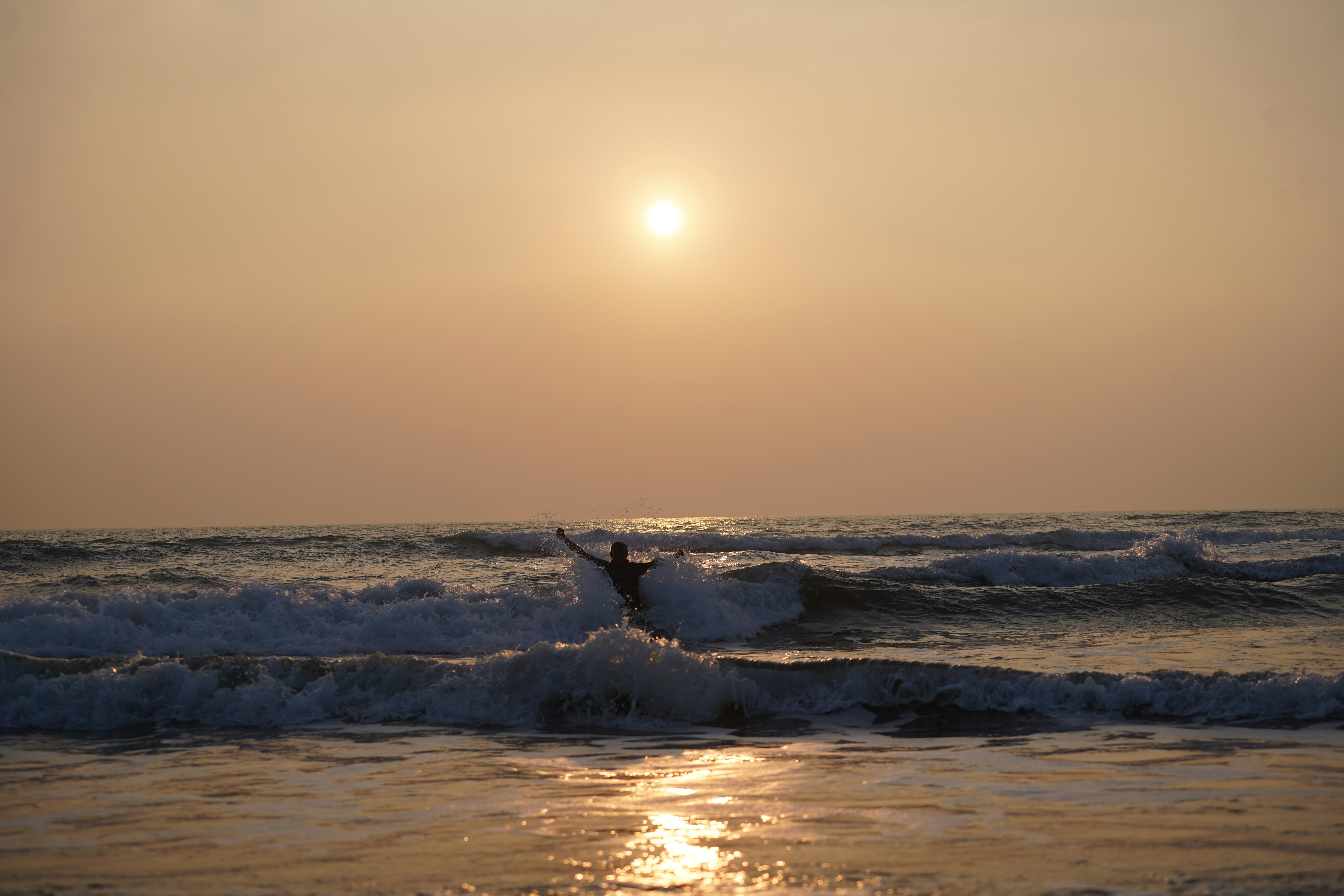 A lone surfer balances on a wave at sunset, with warm horizon light reflecting off the water. The scene captures a small human figure amid expansive sea and sky; a tranquil coastal moment.