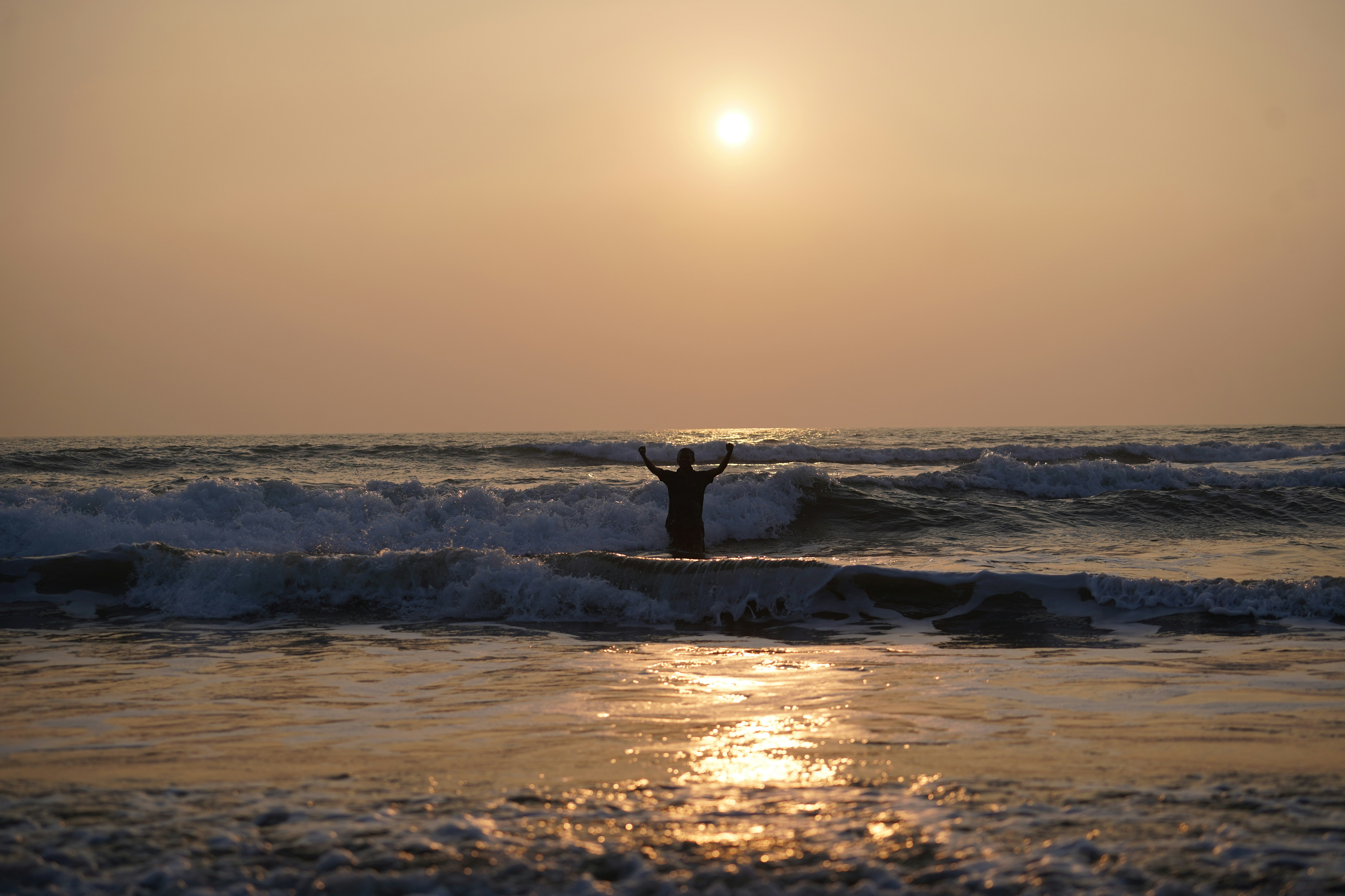 Silhouette of a person with arms raised in the sea at sunset, waves breaking nearby; a tranquil coastal scene with warm tones and reflections on the water. (Photograph)