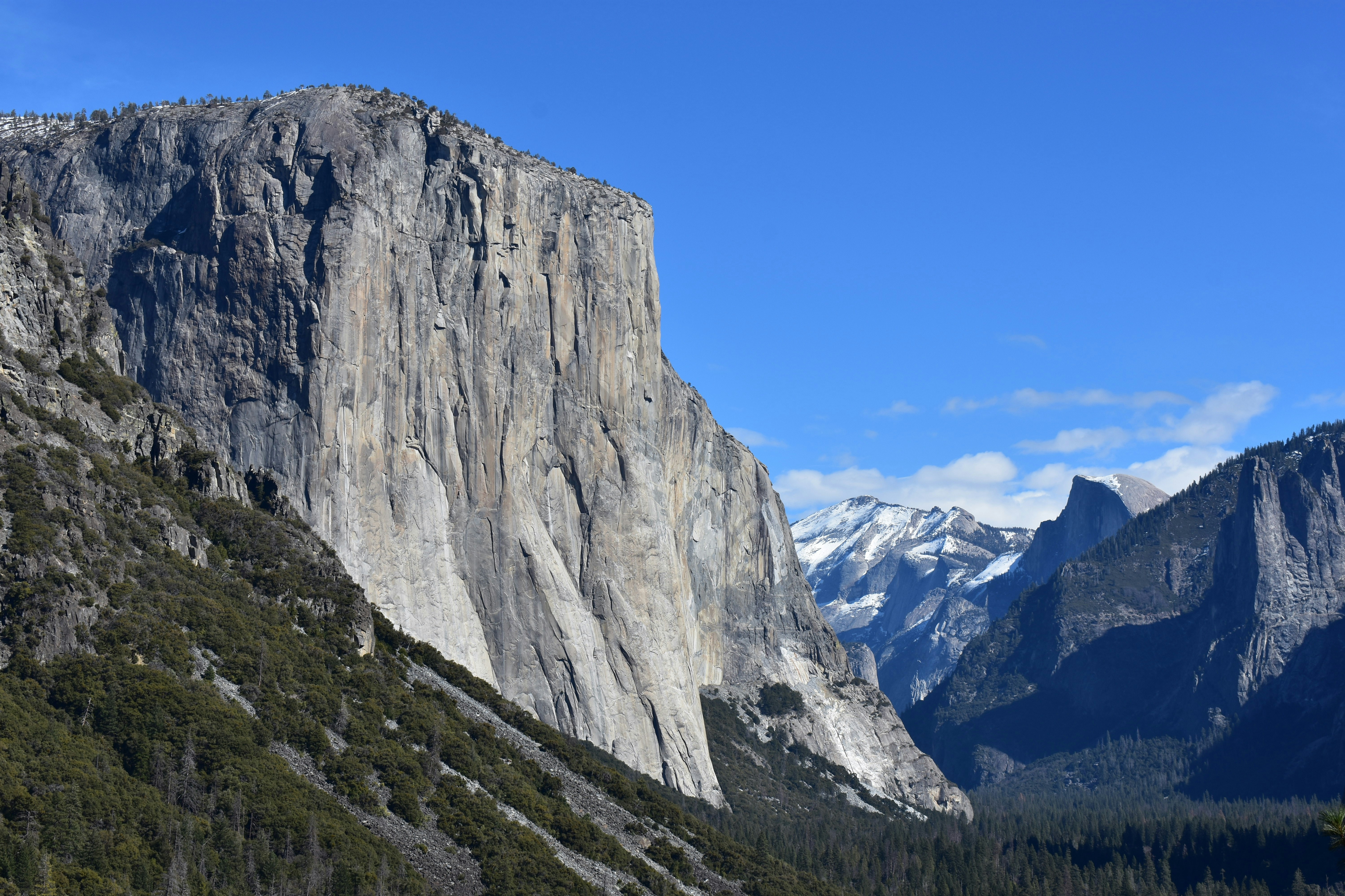 Steep granite cliff over a pine-forested valley framed by distant snow-capped peaks