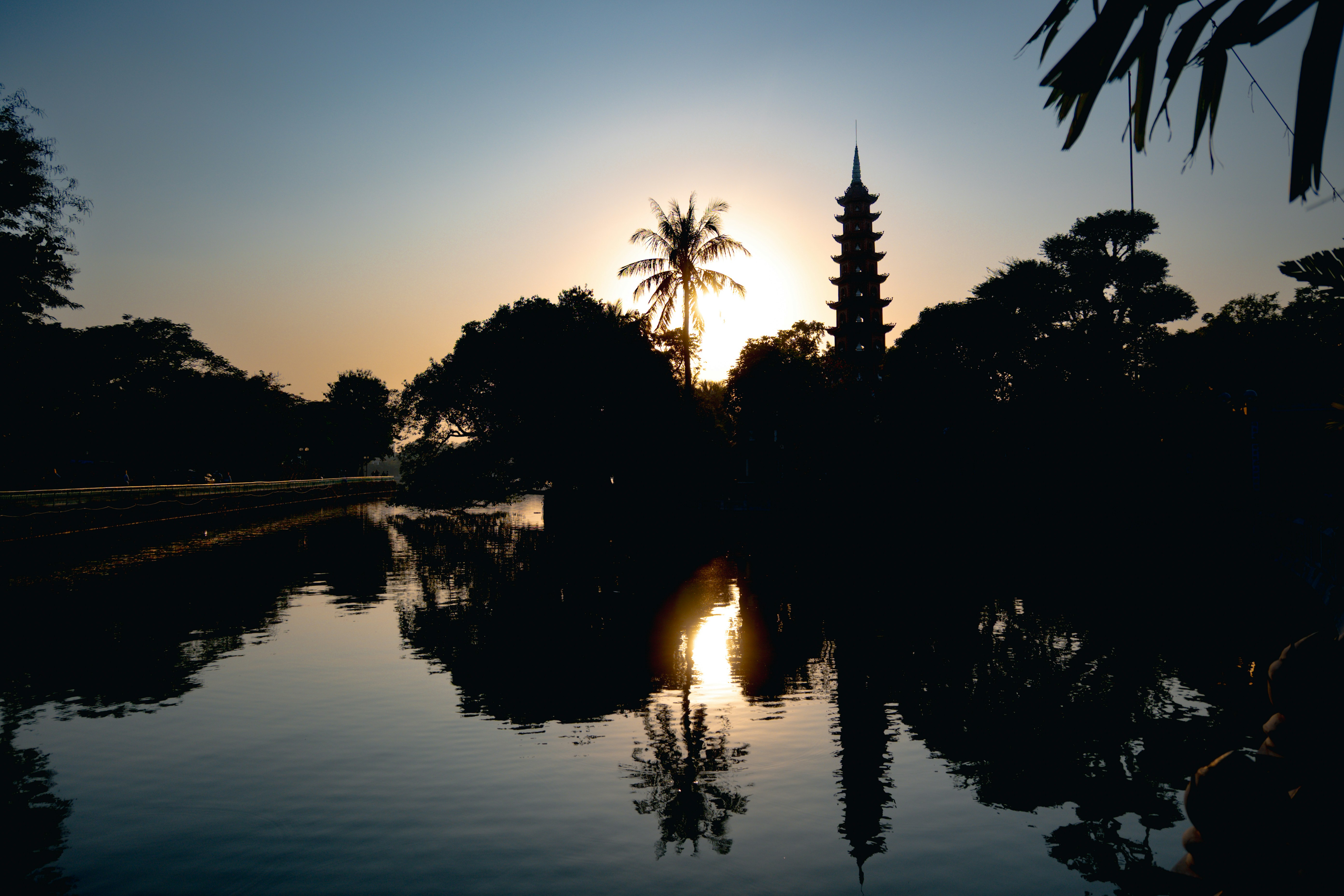 Silhouetted pagoda and palm tree reflected in lake at sunset.