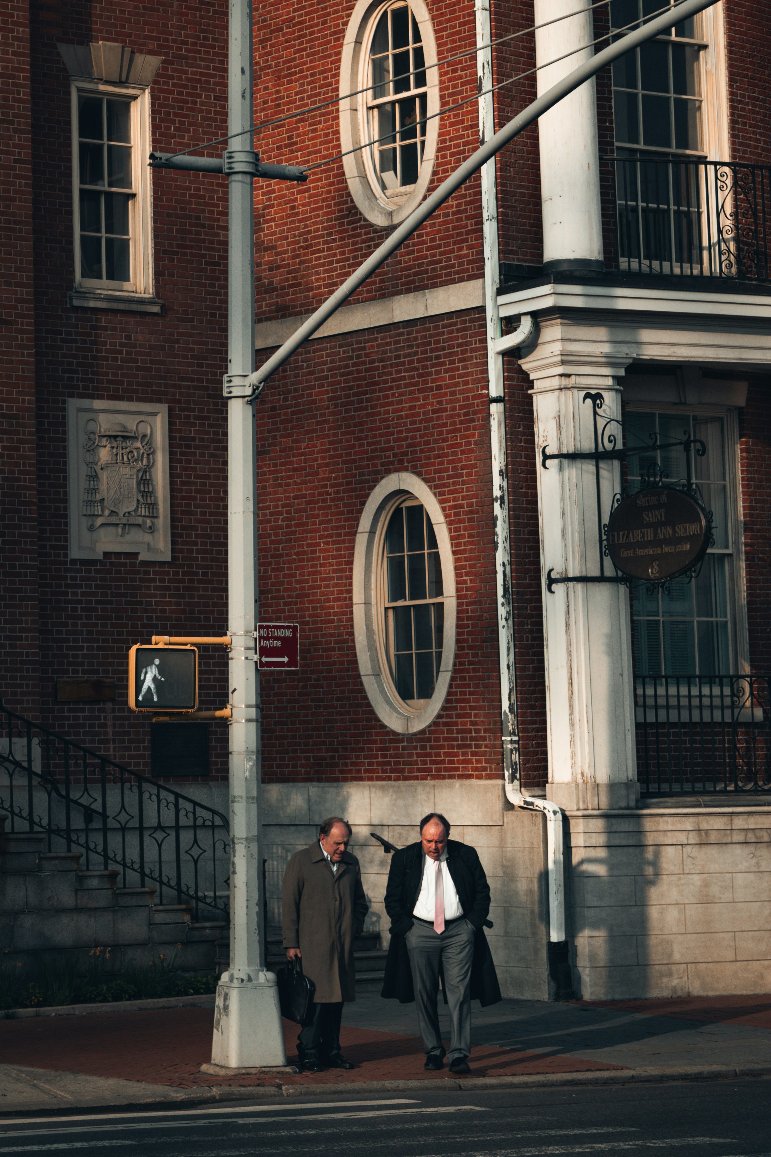 Two men in coats stride past a brick façade beneath a pedestrian crossing sign