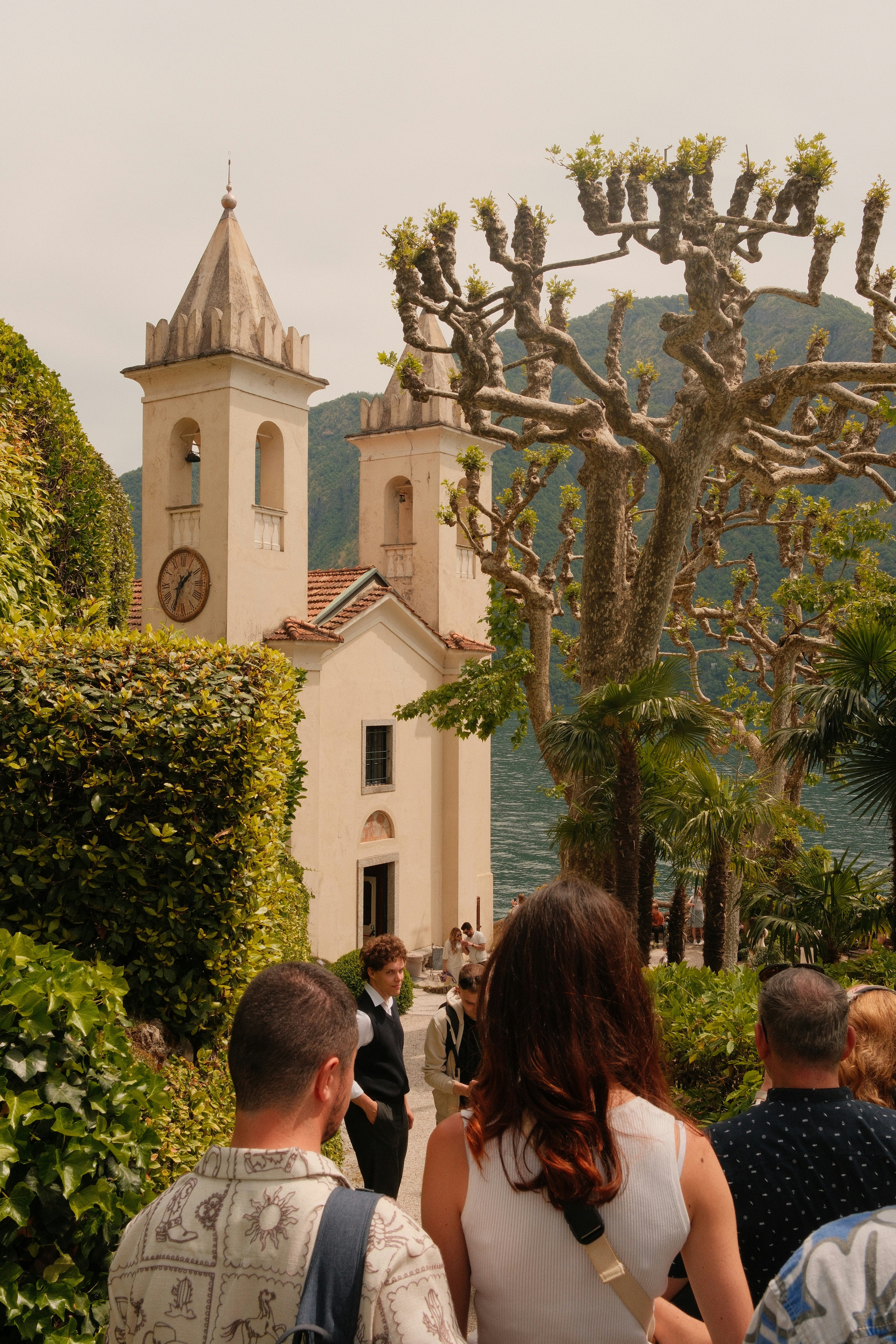 Sunlit Path to the Chapel: A Gathered Crowd En Route by Pruned Trees