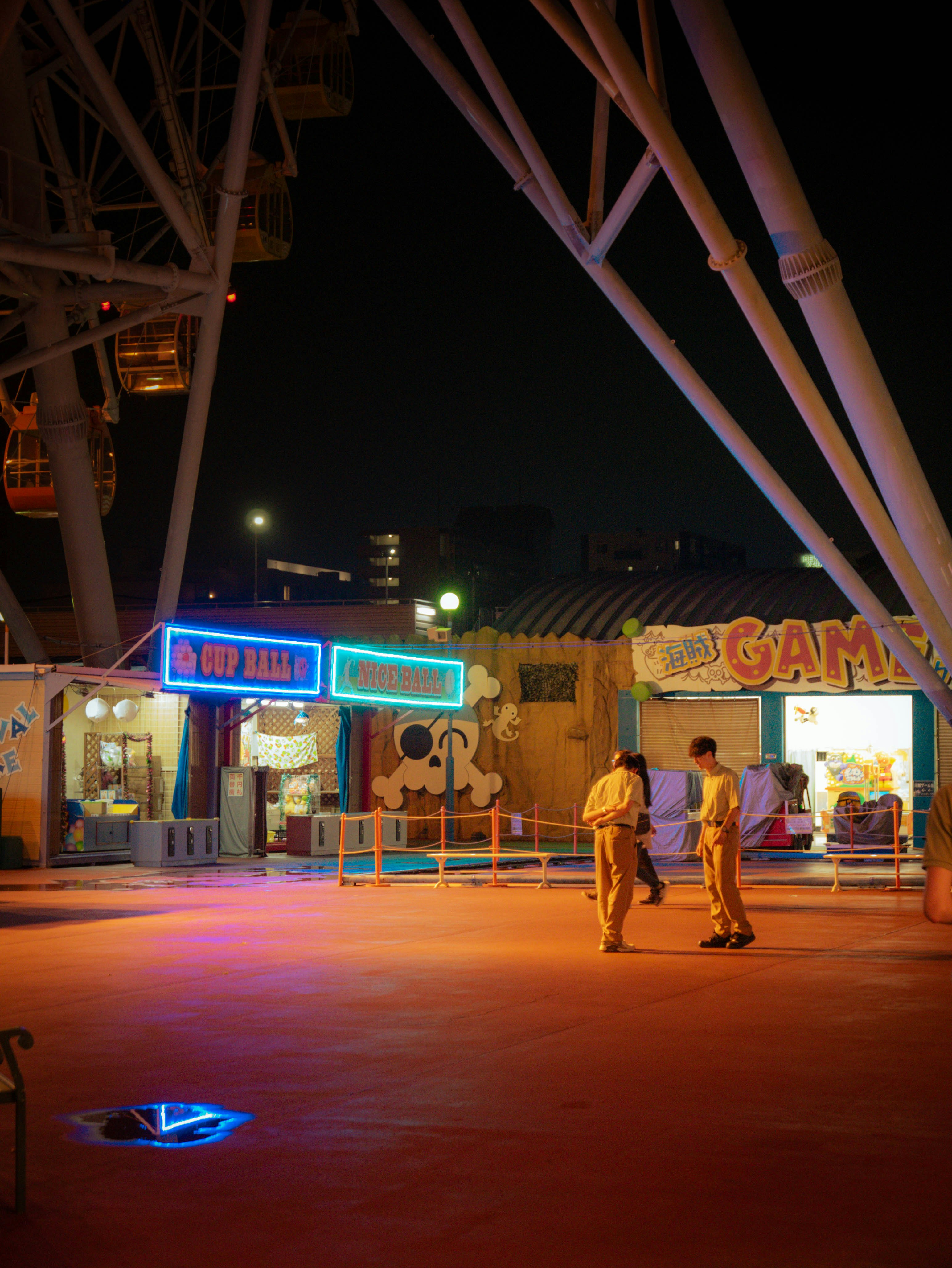 Midnight Carousel of Neon: Two Visitors Pause by the Game Stalls