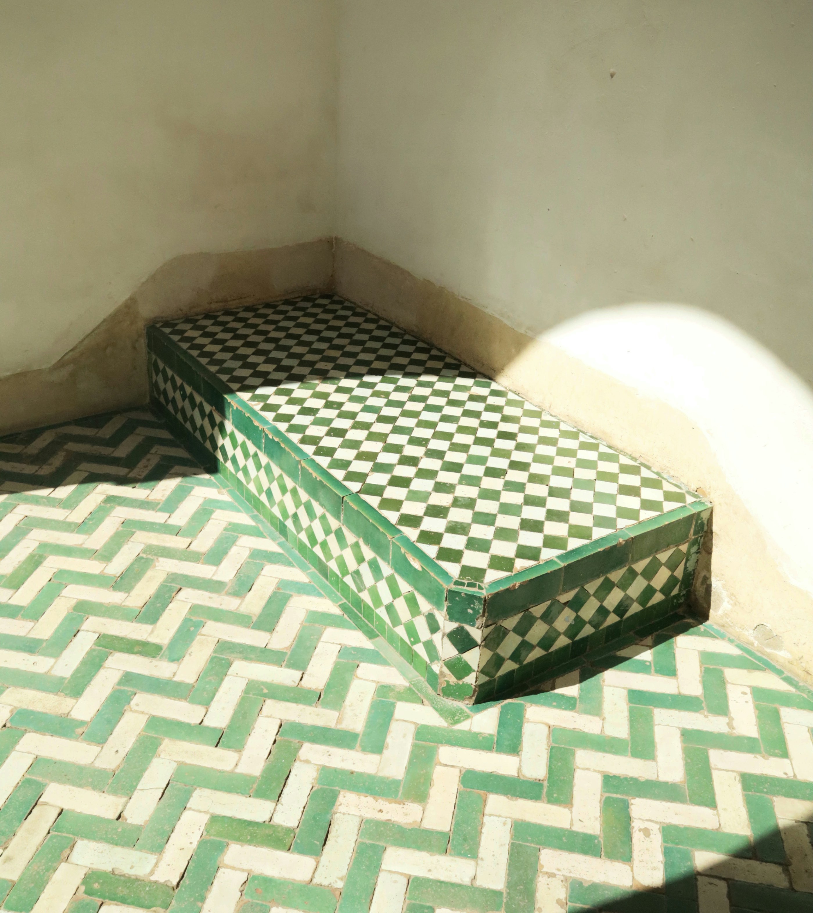A sunlit checker-pattern tiled step sits against a pale corner wall, with bold green-and-white geometric flooring extending outward.