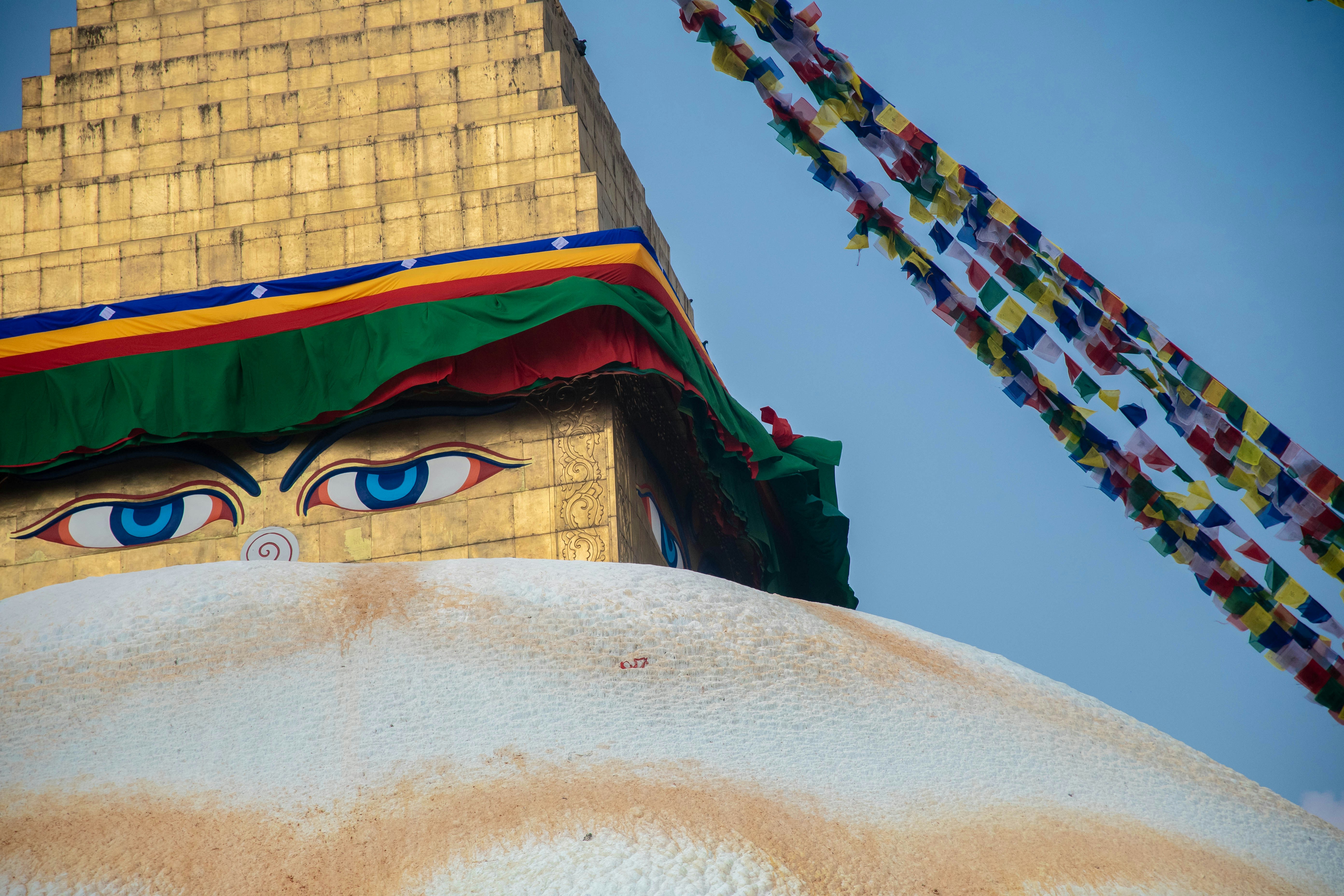 Golden stupa with painted eyes and colorful prayer flags fringing the roof, set against a clear blue sky; the scene emphasizes spiritual symbolism and cultural ritual.