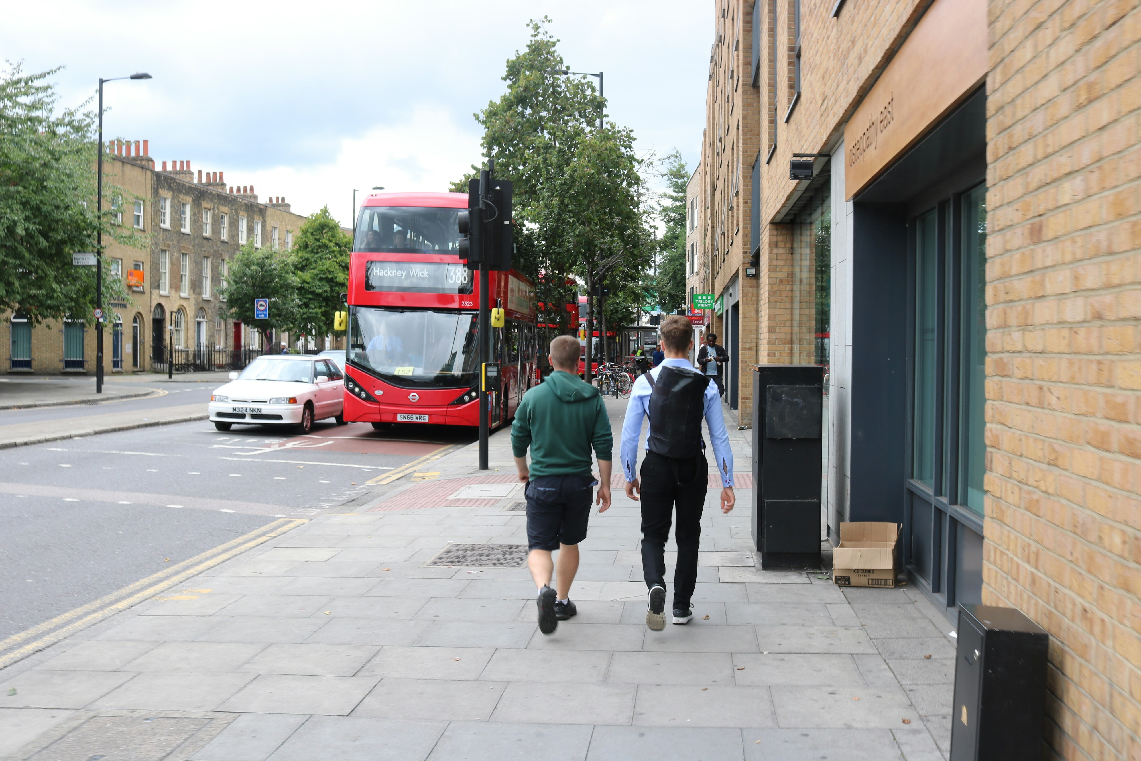Two pedestrians stride past a storefront as a red double-decker bus glides by on a city street