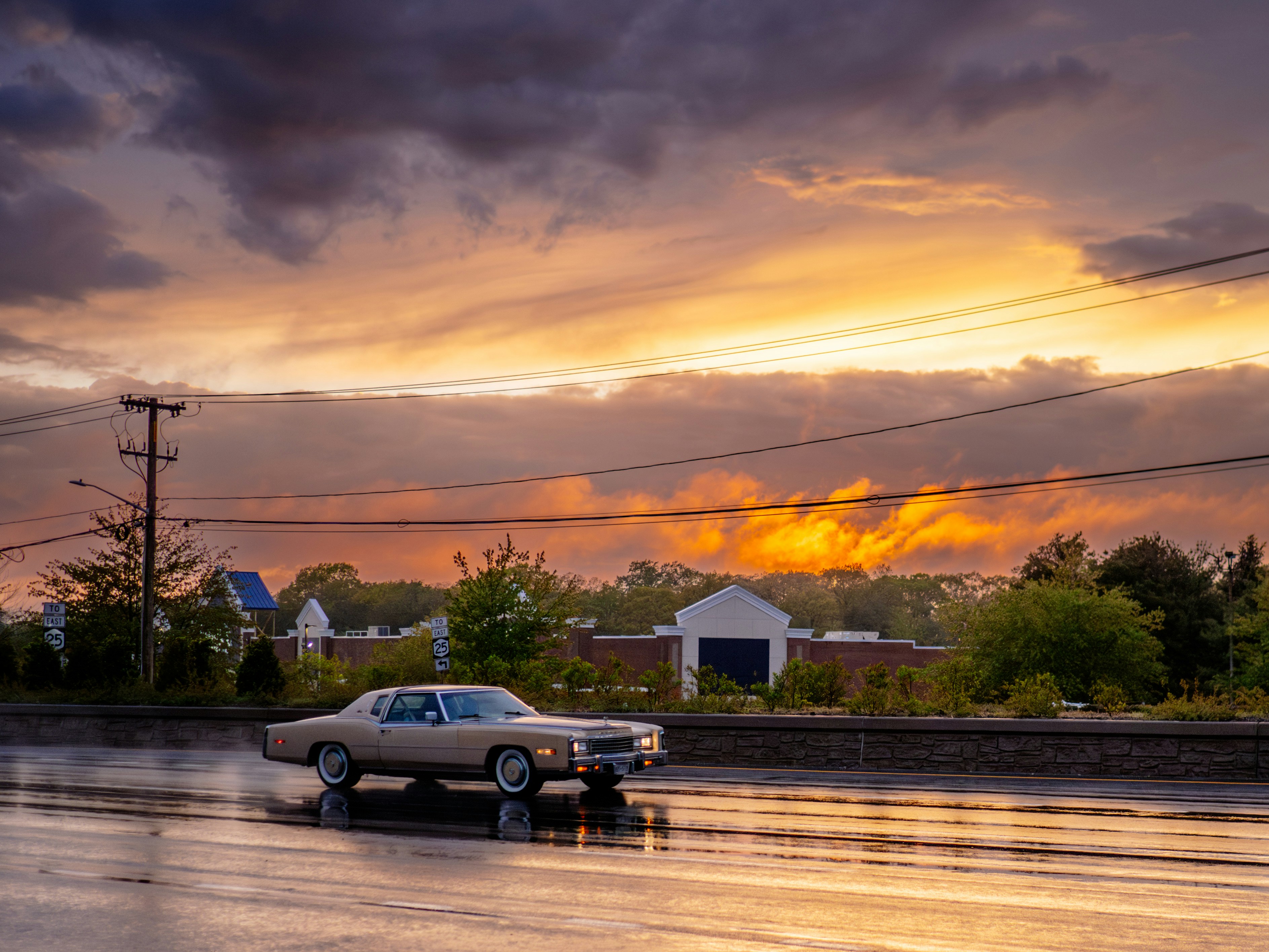 Drenched Streetlight Sunset: A lone vintage car glides through a reflective road as evening clouds burn above