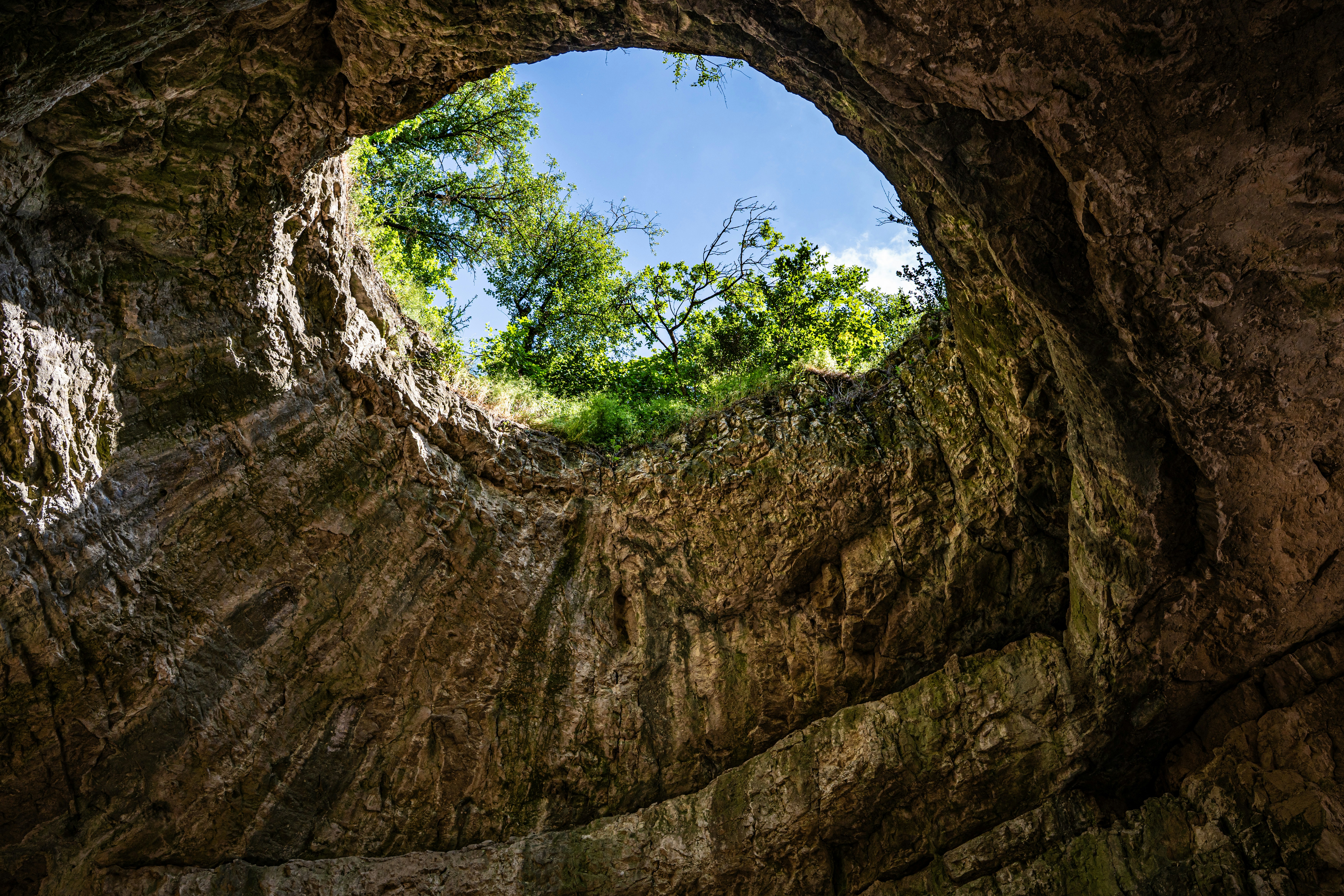 Circular cave opening framing a blue sky and green canopy; rugged rock walls enclose a natural aperture.