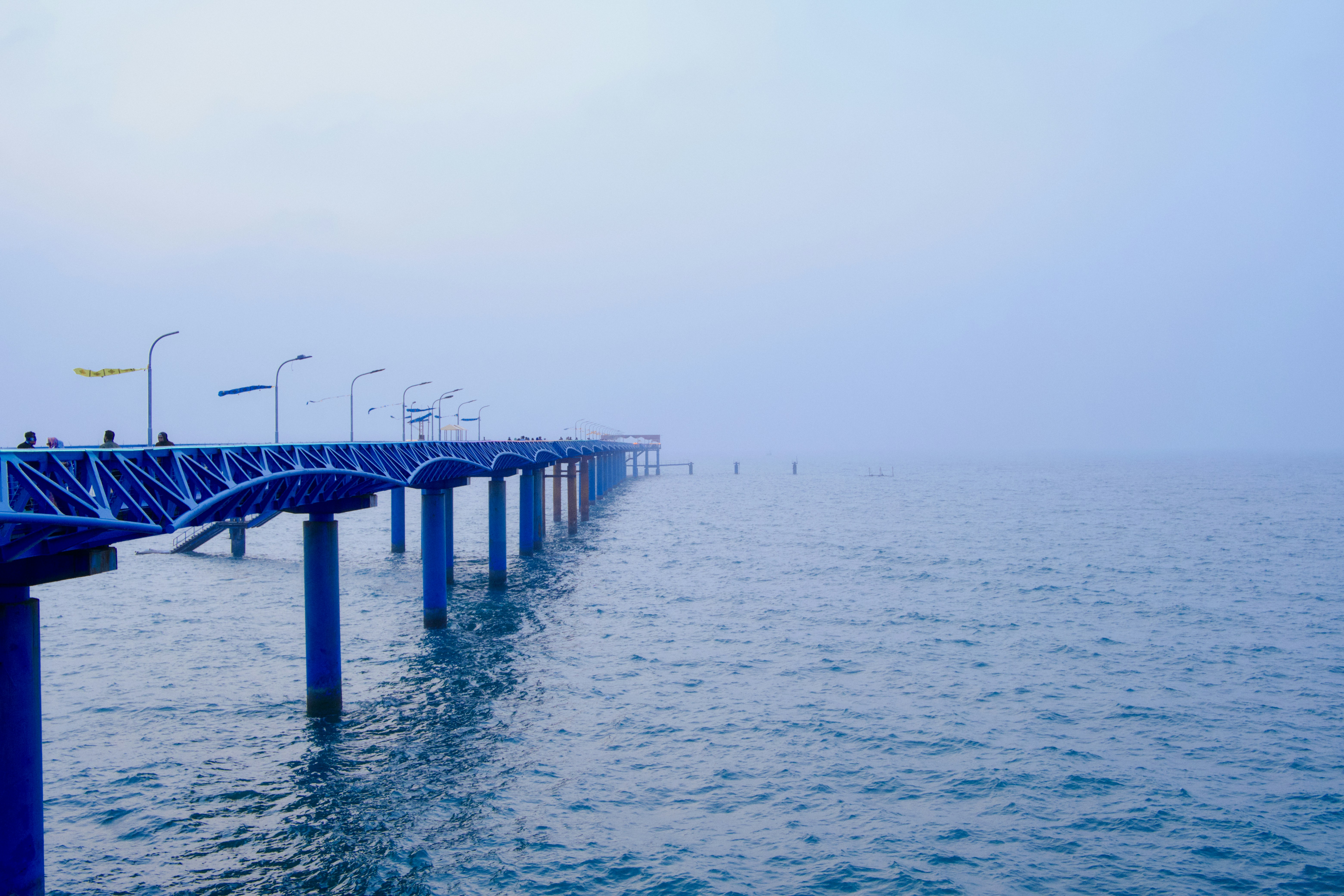 Blue pier stretches into a calm, misty sea with soft lighting and a hazy horizon, emphasizing tranquil seascape architecture.