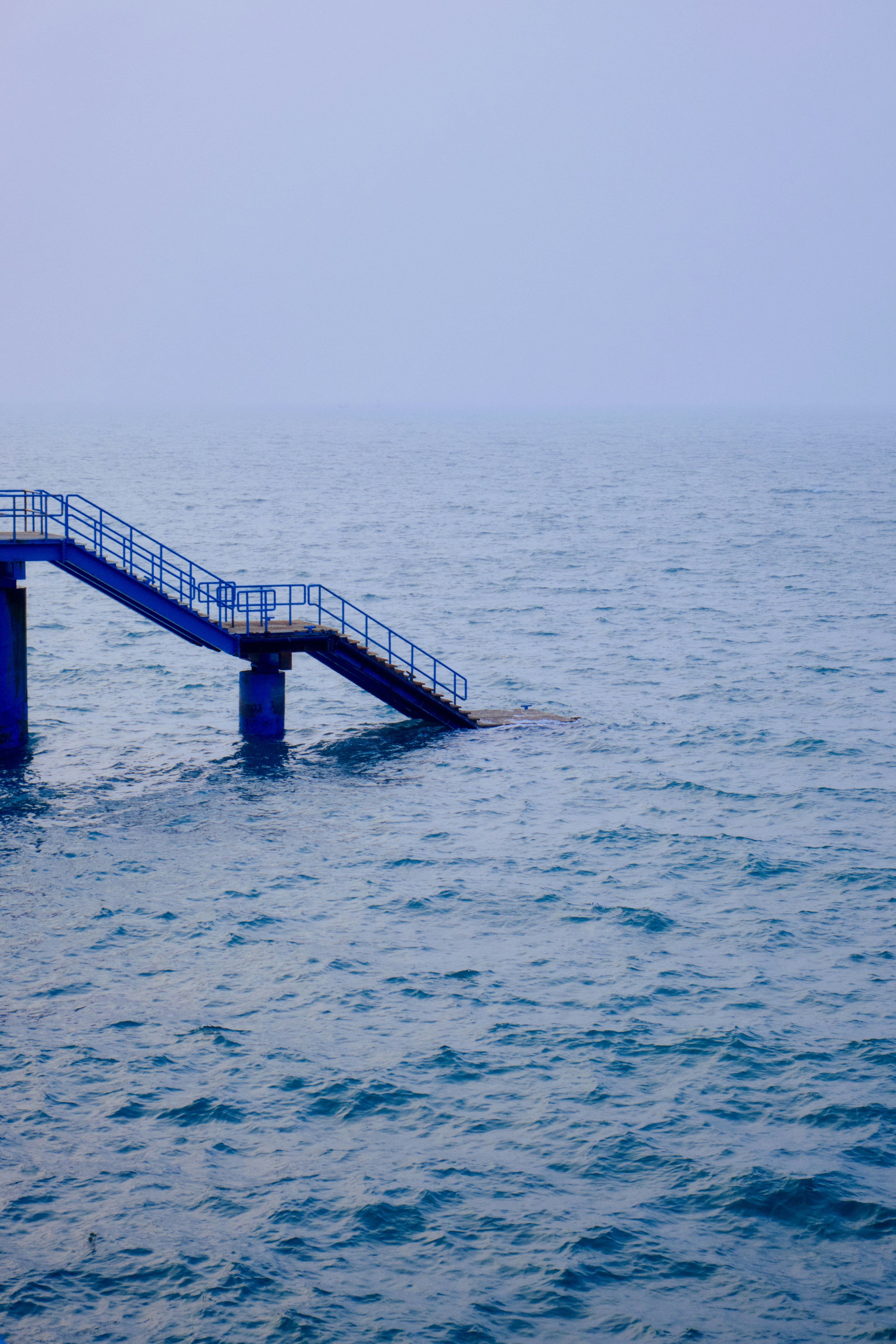 A blue seaside stairway extends into calm ocean waters, with soft horizon and pale sky in the background. The scene emphasizes solitude and the gentle gradient of blue tones.