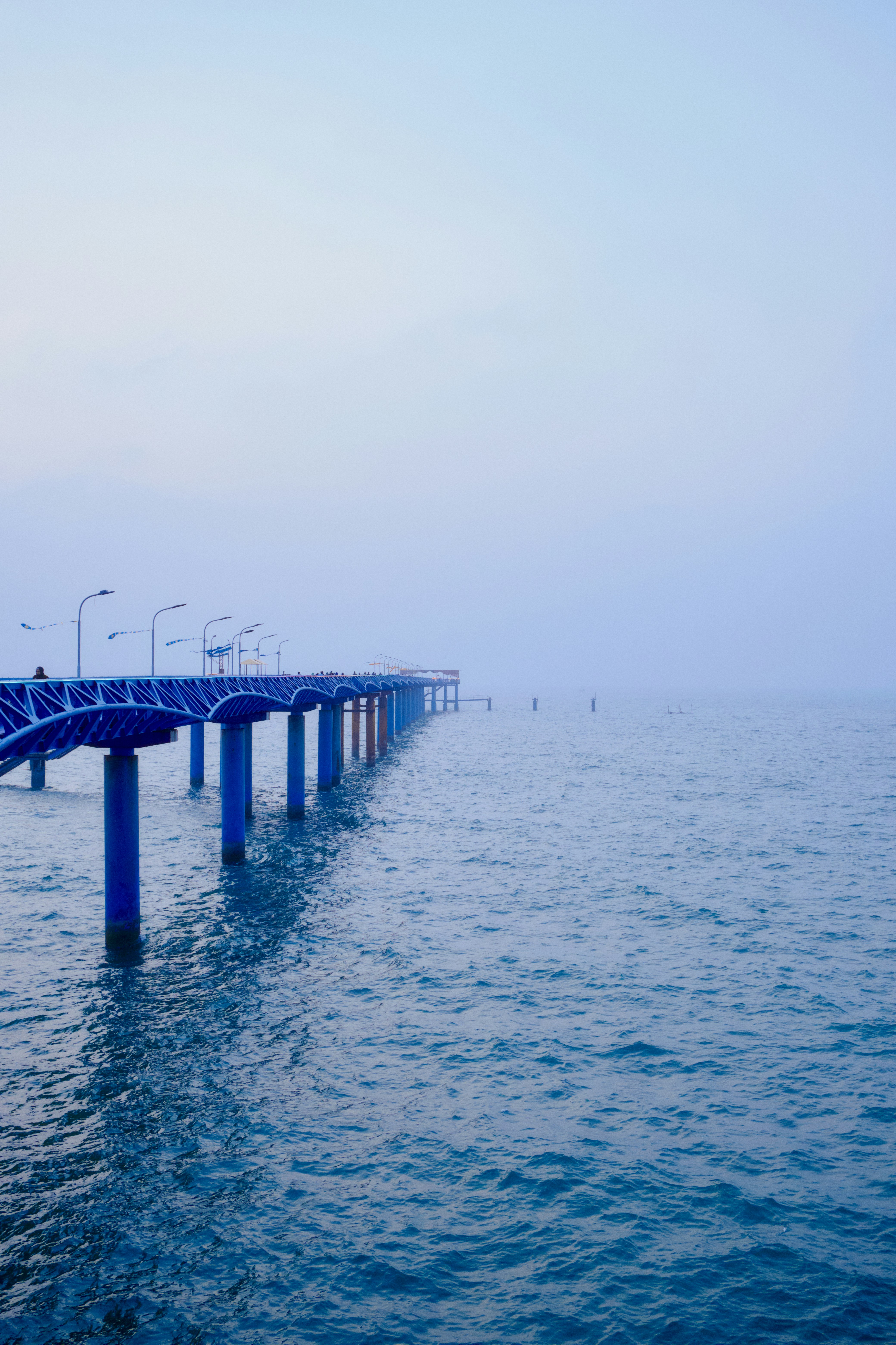 Blue pier extends into calm sea under pale sky, with evenly spaced lamps along the railing and weathered supports fading toward the horizon.