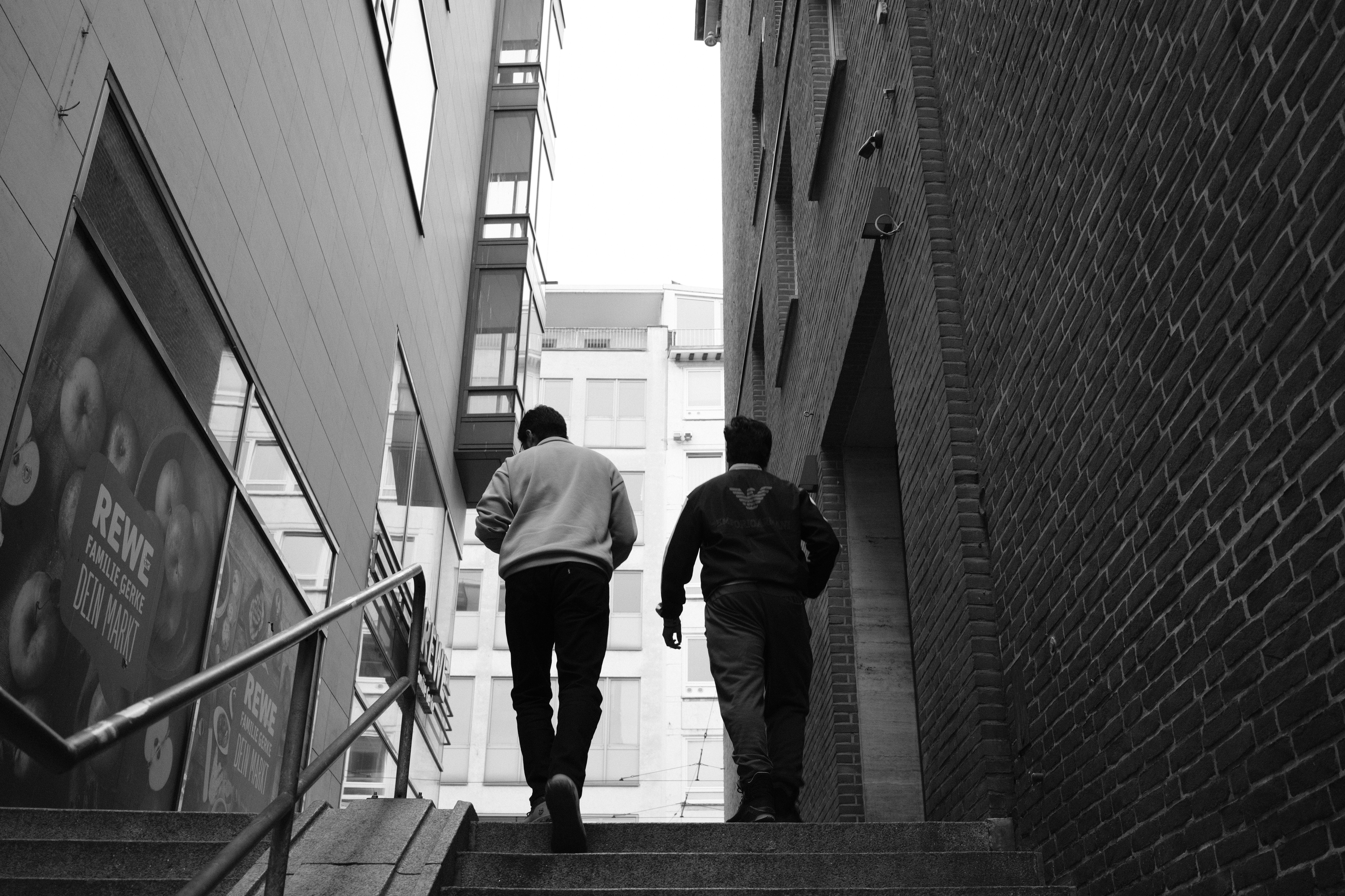Two Figures Ascend the Urban Stairwell Toward a Bright Gap