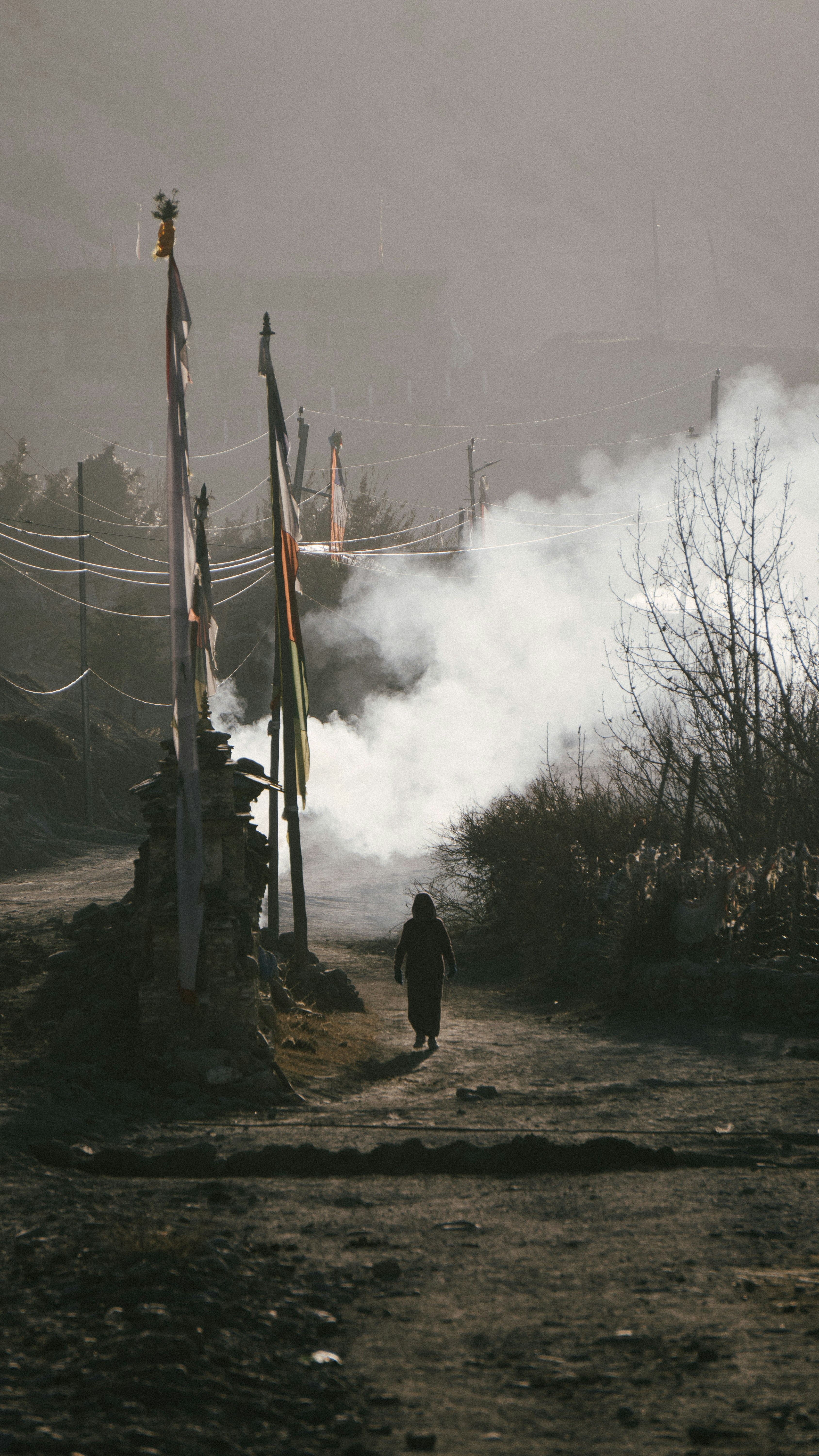 Whispering Path: A lone figure and prayer flags along a smoke-veiled road