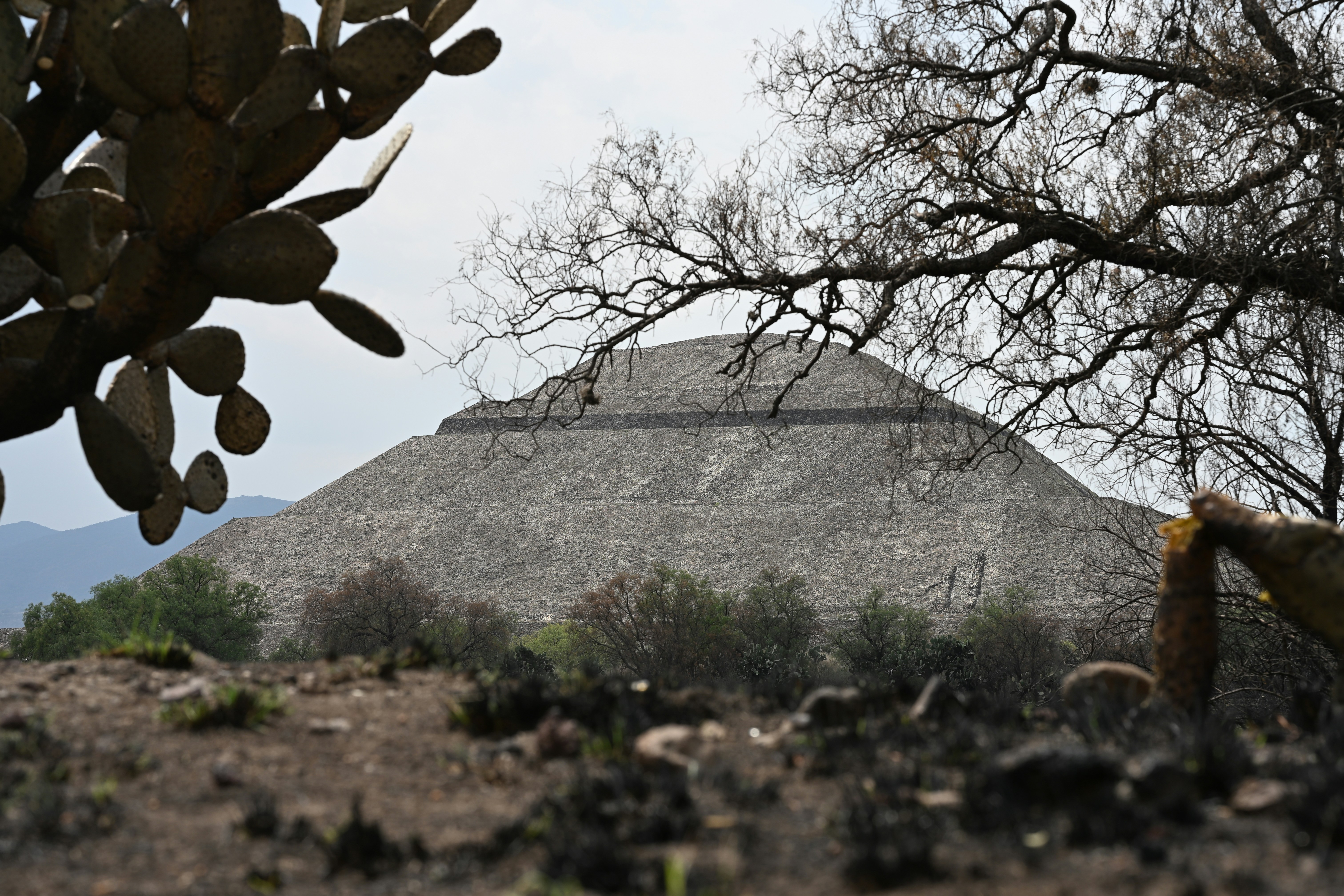Desert hillside with a stepped, pyramidal hill framed by sparse vegetation and a distant blue sky. The scene emphasizes the lone, monumental mound against a rugged foreground.