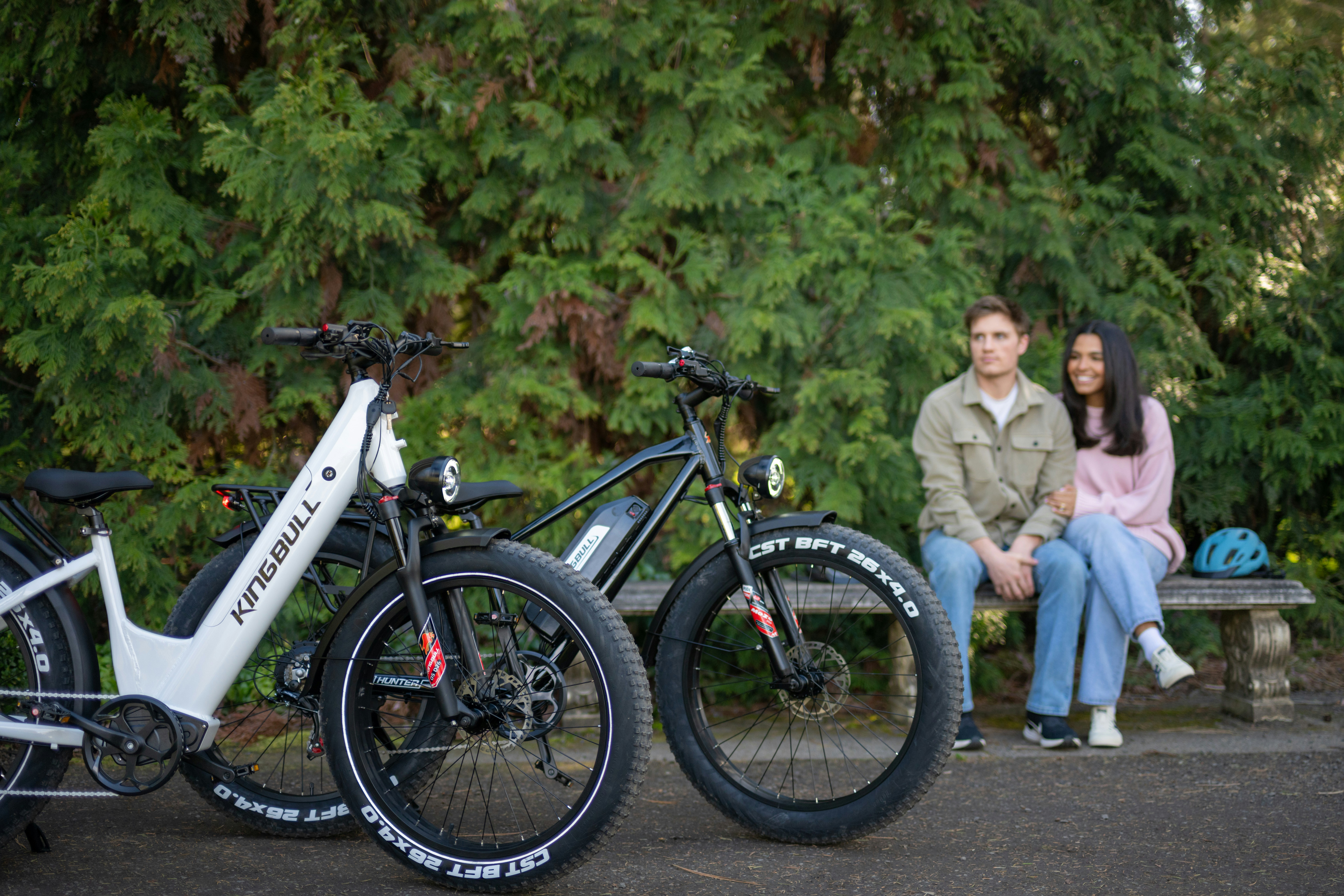 Two riders pause beside a pair of electric bikes under a leafy backdrop