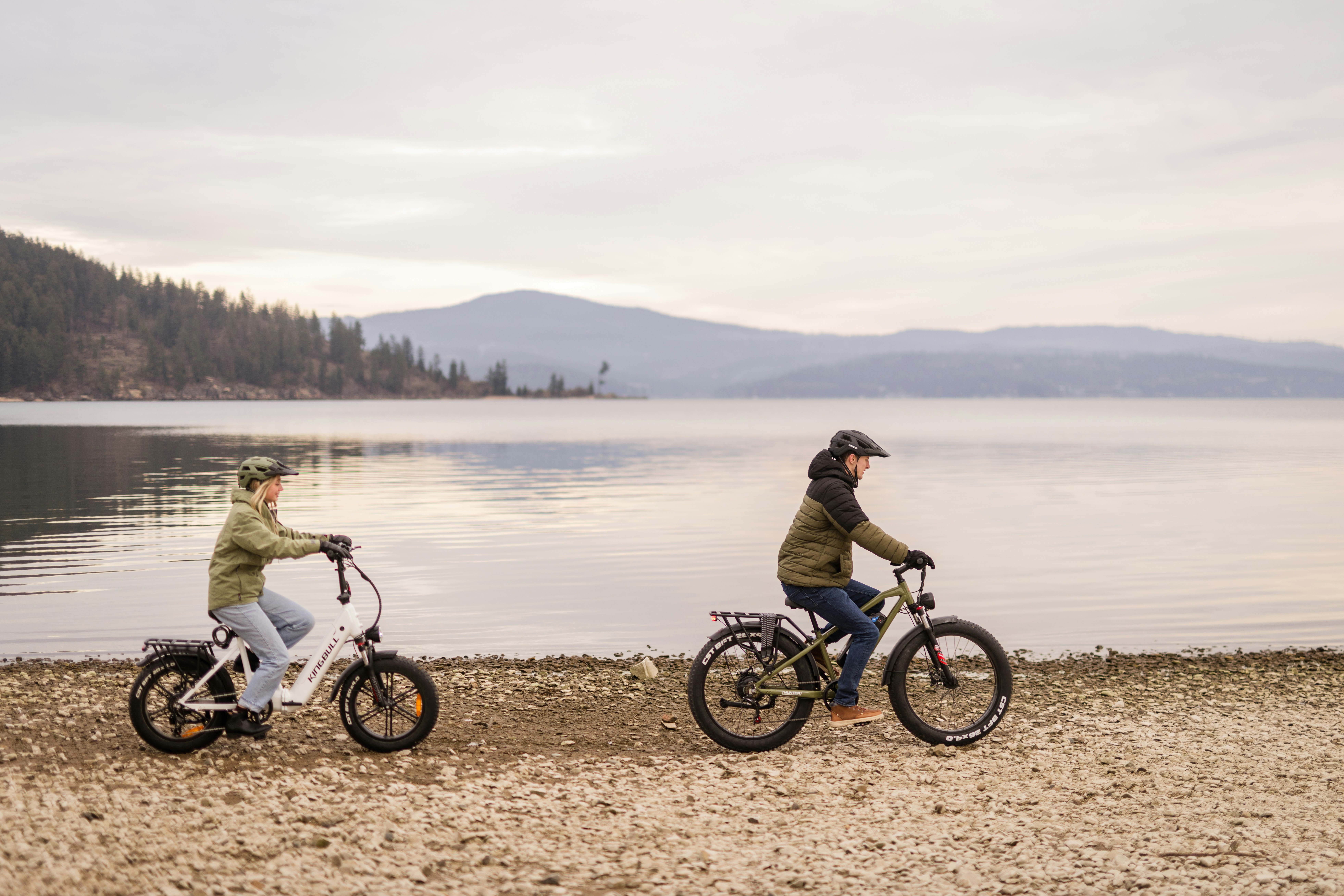 Two riders along the pebbled shore with calm lake and distant mountains
