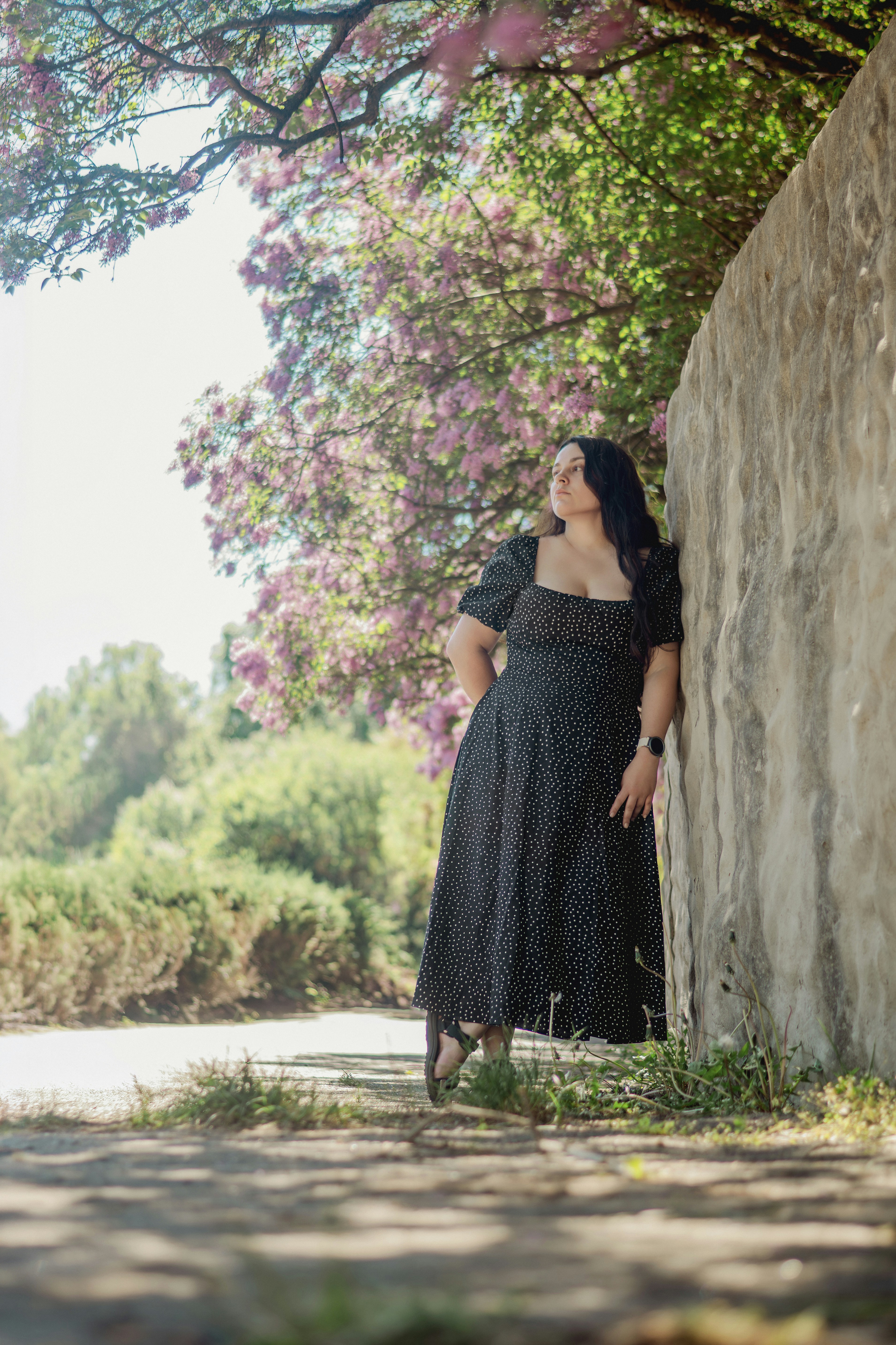 Woman in a dotted black dress leans against a sunlit stone wall beneath blooming pink blossoms