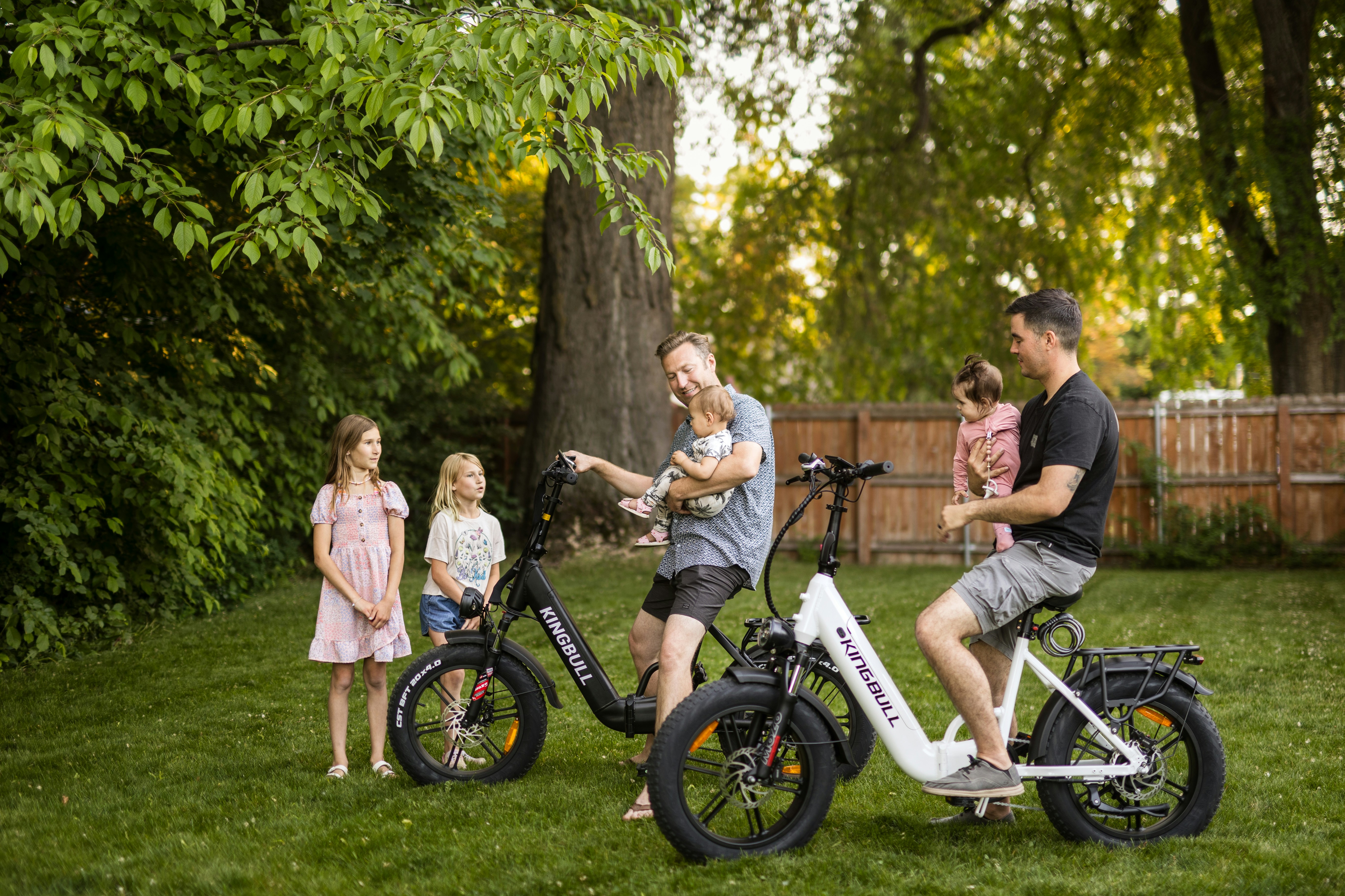 Family ride-time: two electric bikes parked as parents cradle toddlers while kids observe in a sunlit park